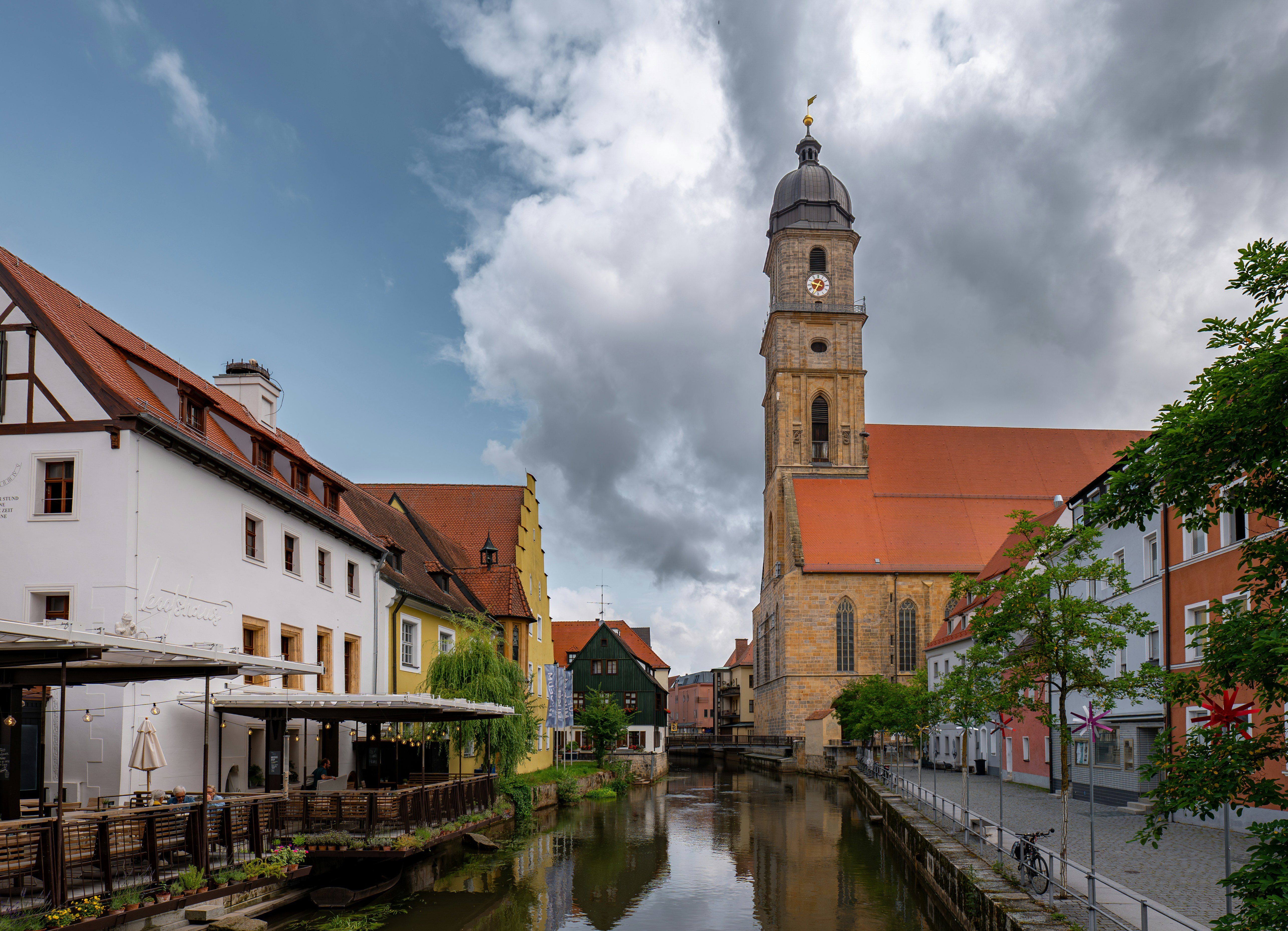 Historic buildings lining a tranquil canal, with a prominent clock tower rising above the scene. Lush greenery adds a touch of nature to the urban landscape.