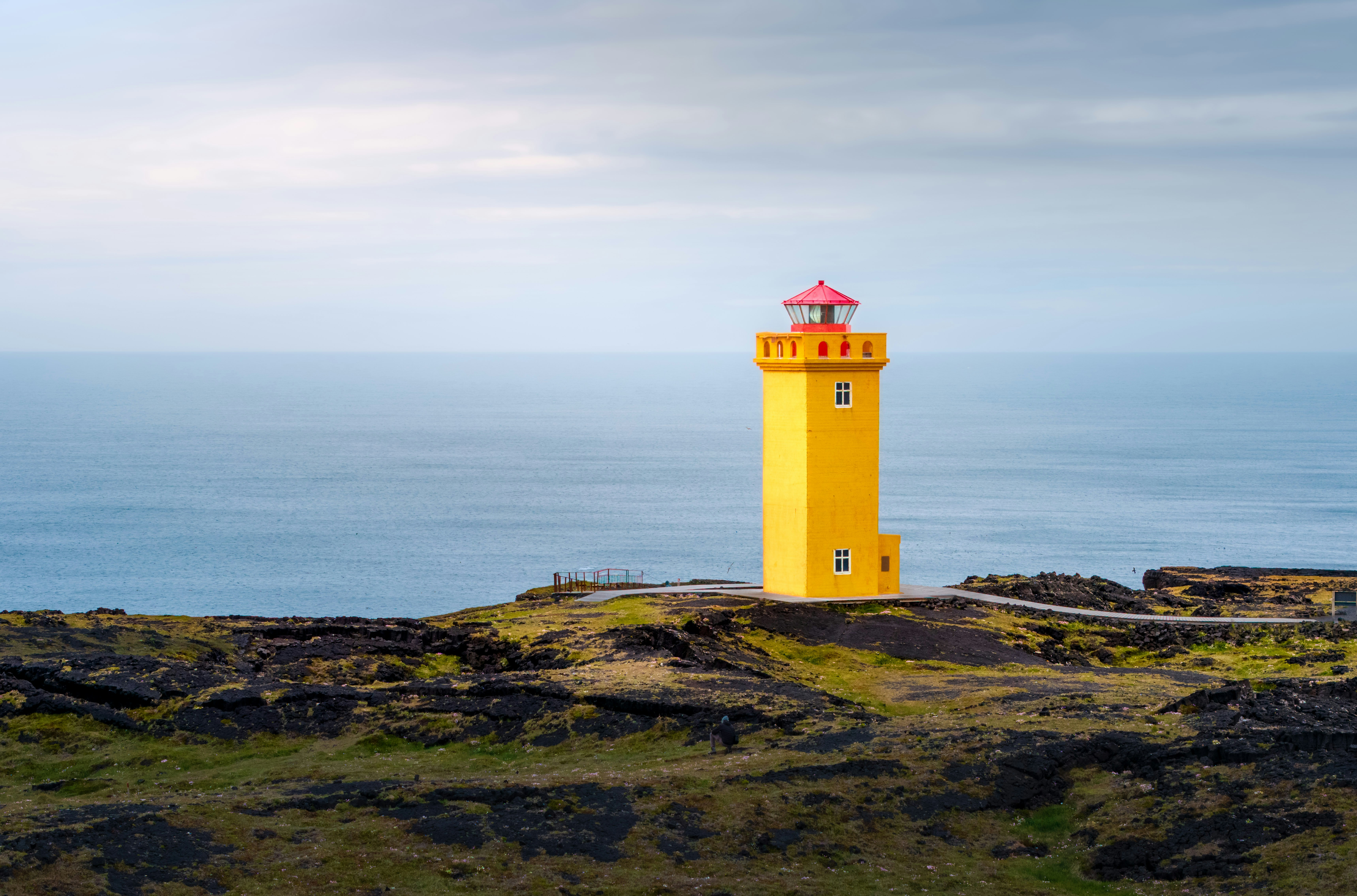 Svörtuloft Lighthouse’s bright yellow tower stands out against rugged volcanic cliffs as the vast Atlantic Ocean stretches to the horizon in a dramatic Icelandic seascape. | A bright yellow lighthouse stands overlooking the sea.