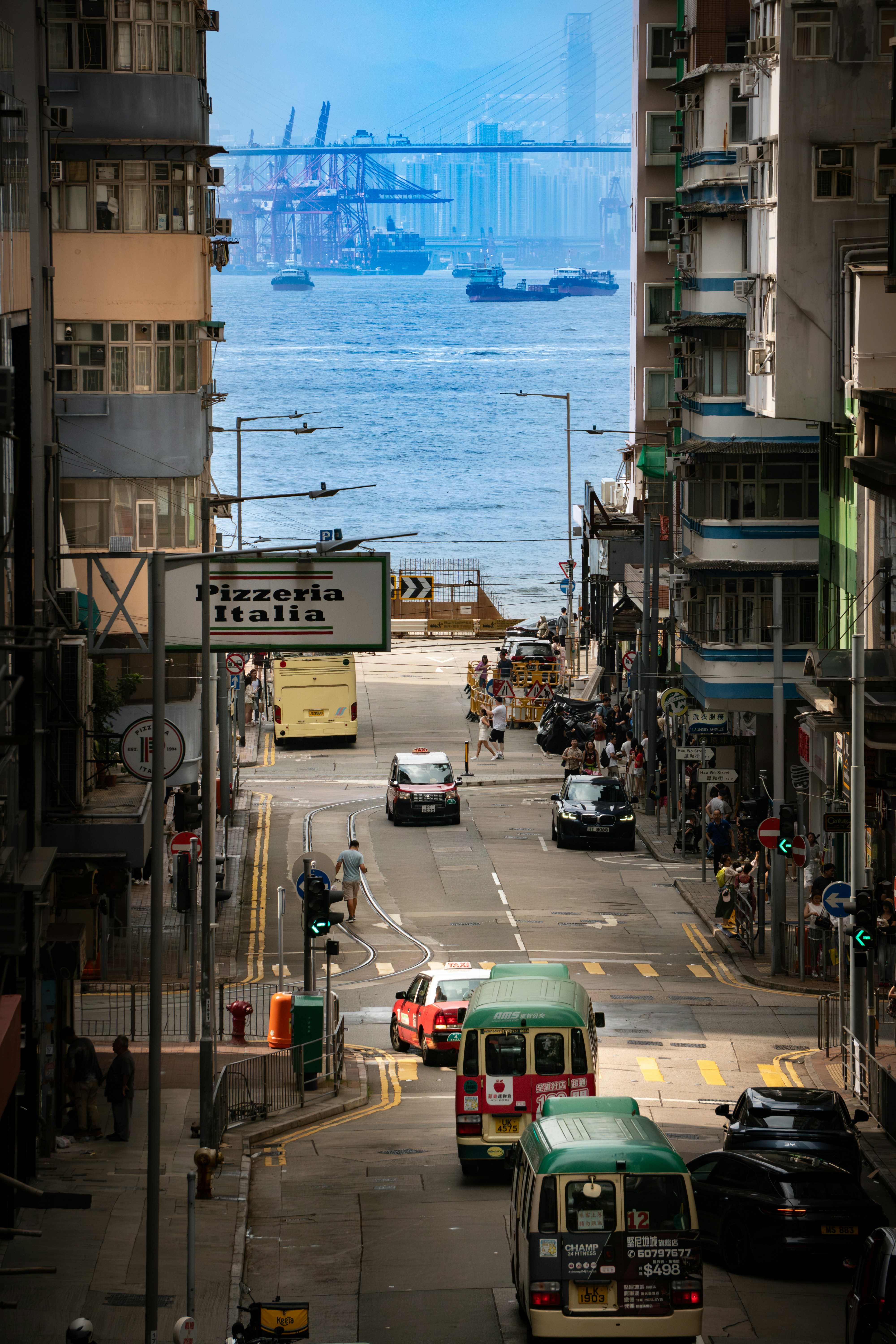 Busy street scene in Hong Kong showcasing vehicles and pedestrians with a backdrop of the harbor and distant cranes. Pizzeria Italia sign adds a local touch.