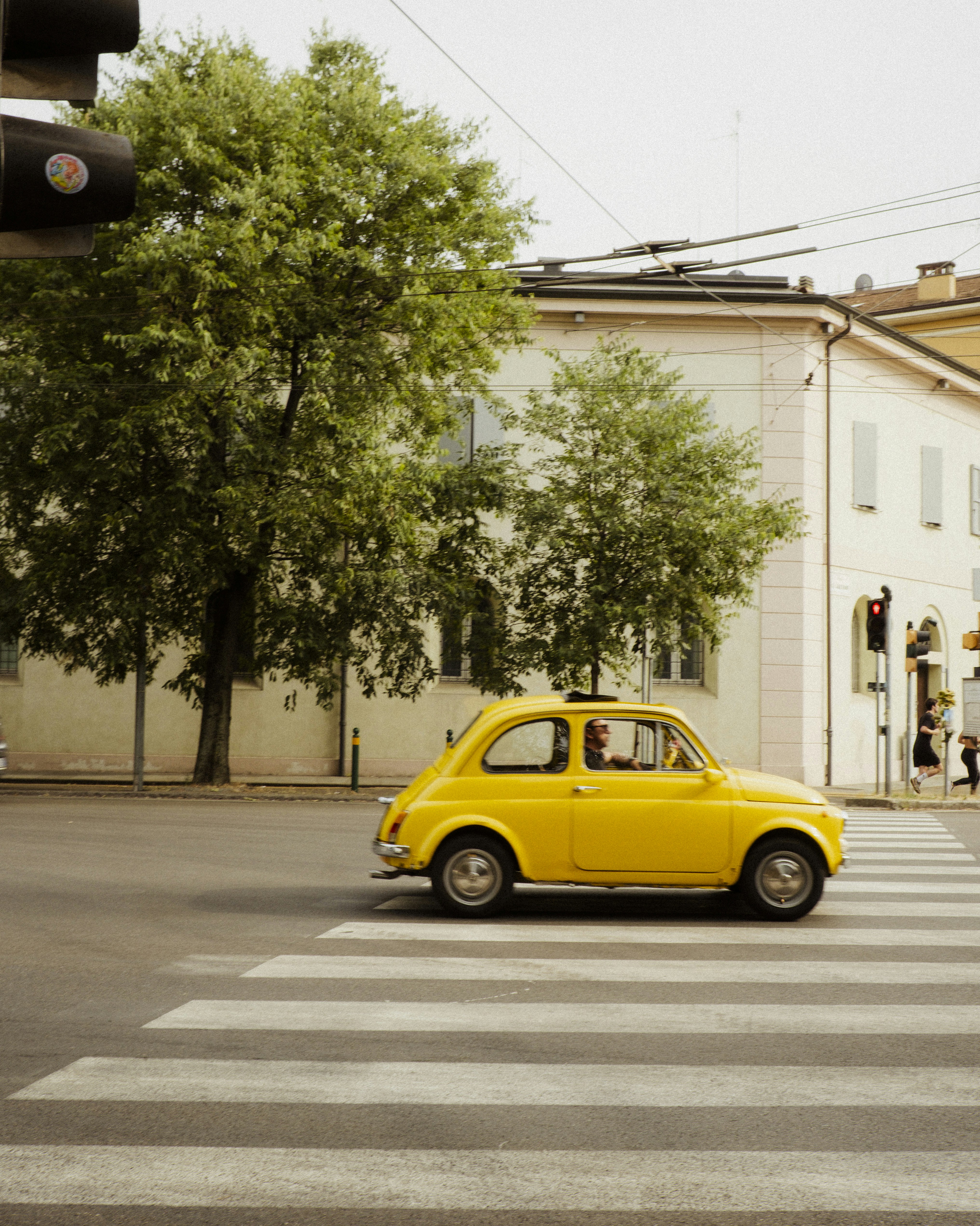 Classic yellow car cruising past a crosswalk with pedestrians in a charming urban setting.