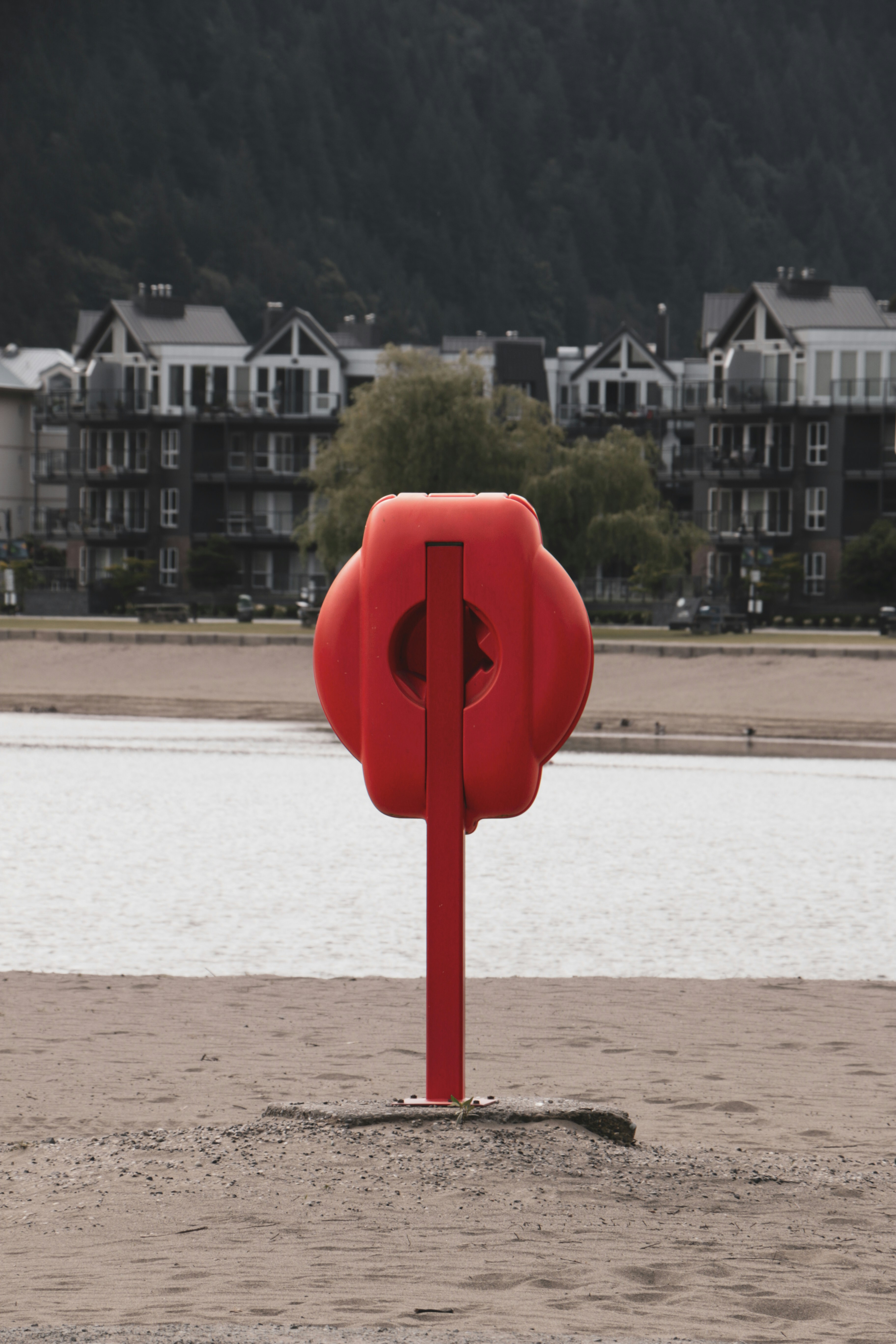 Bright red life preserver mounted on a post, set against a backdrop of modern buildings and a calm waterway.