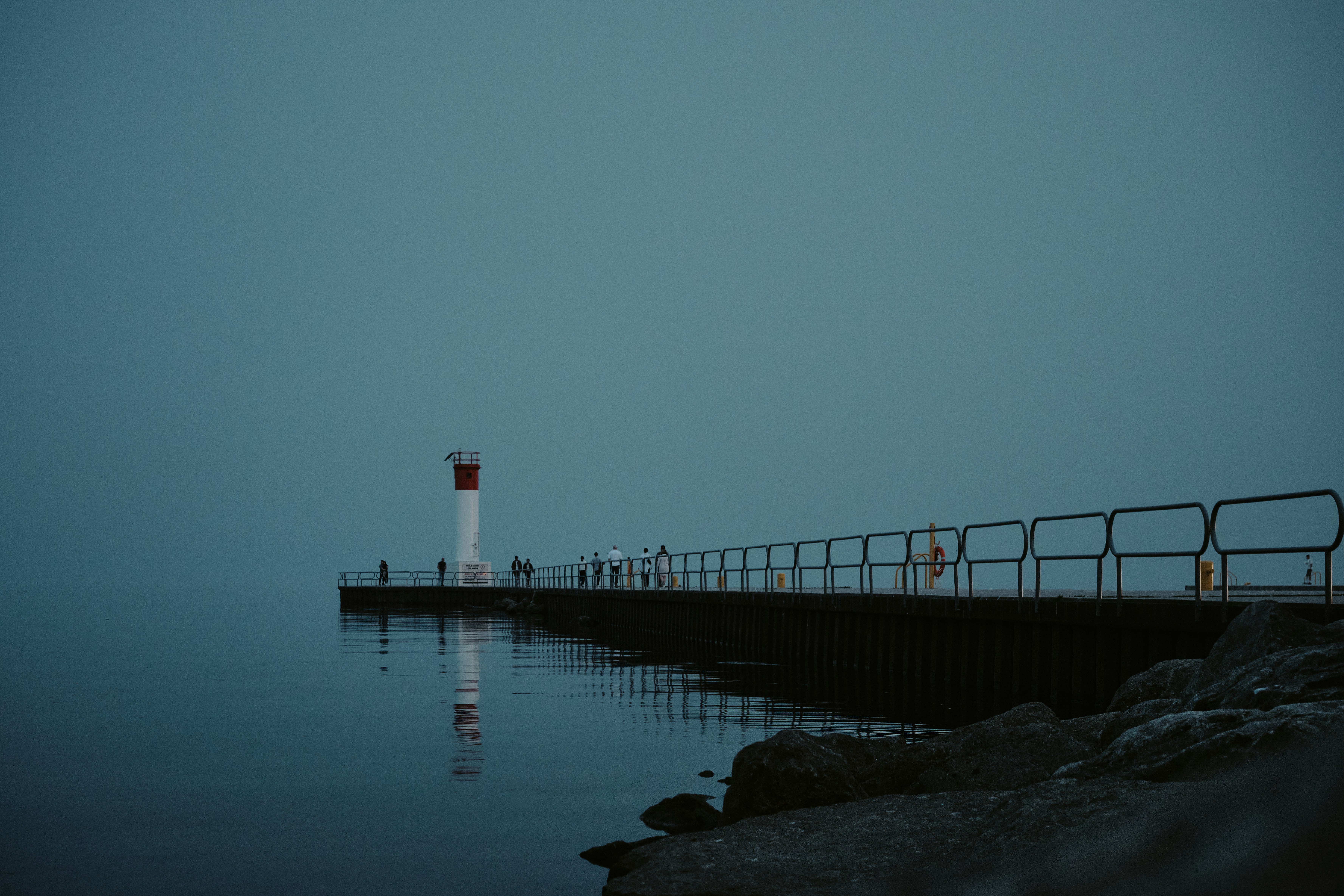 A calm evening settles over the lake as silhouettes gather near a distant lighthouse. The cool dusk light wraps the pier and water in soft blues, creating a sense of peace and quiet solitude. | Lighthouse stands at the end of a pier.
