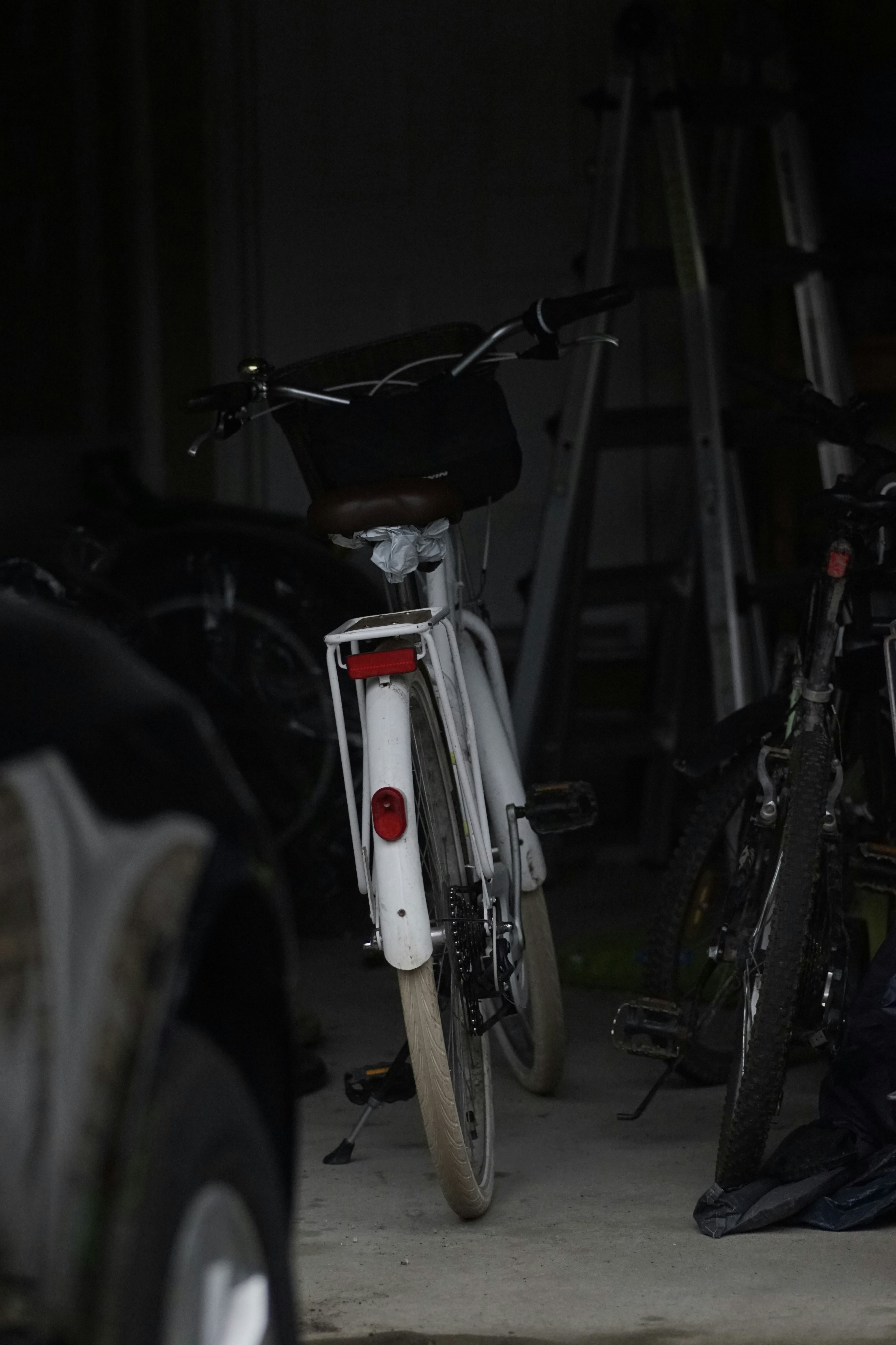 A white bicycle rests in a dimly lit garage, surrounded by various tools and other bicycles. The scene conveys a sense of stillness and forgotten moments.