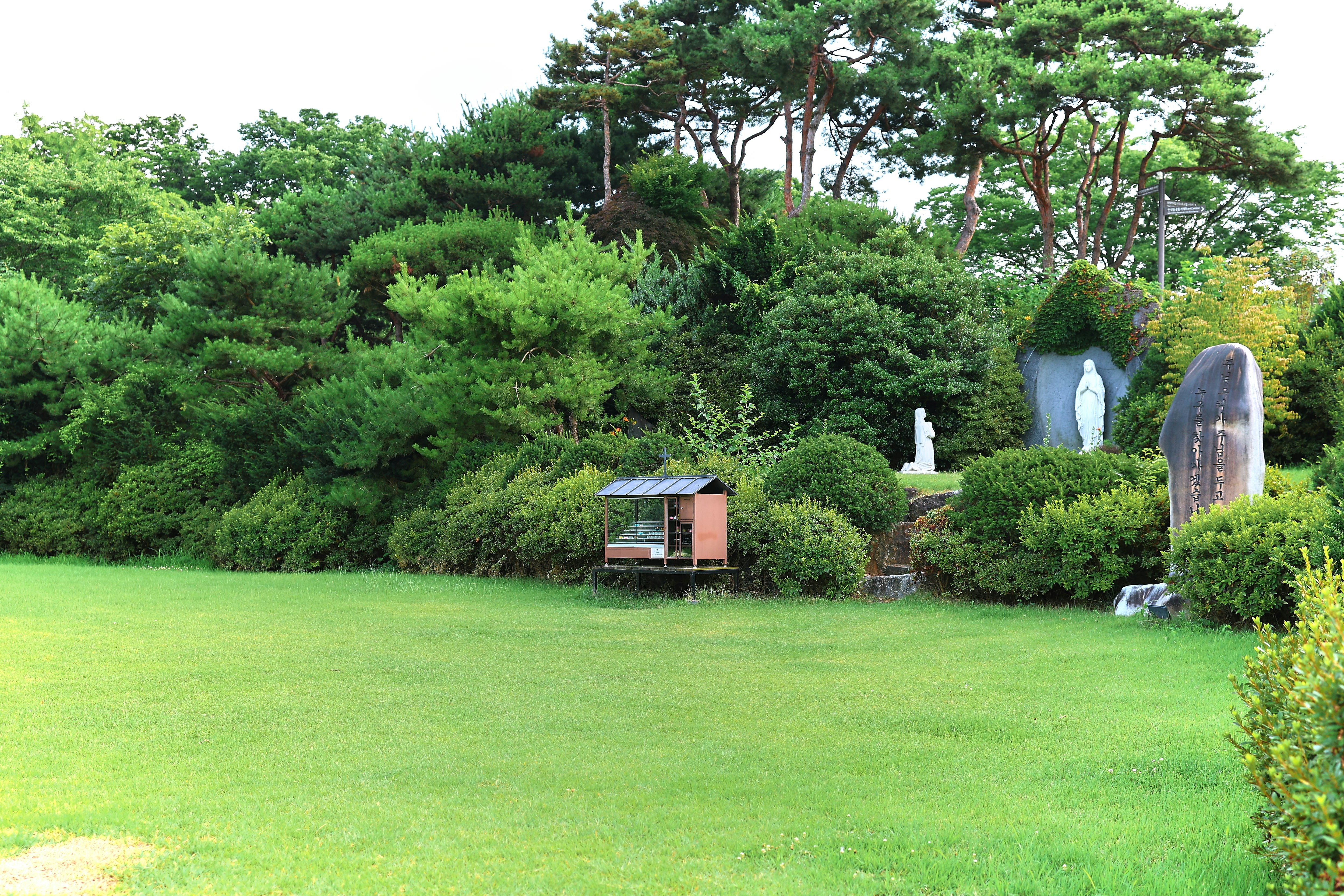 Lush greenery surrounds a serene garden space featuring a statue and a small shelter. The tranquil setting invites reflection and peace.