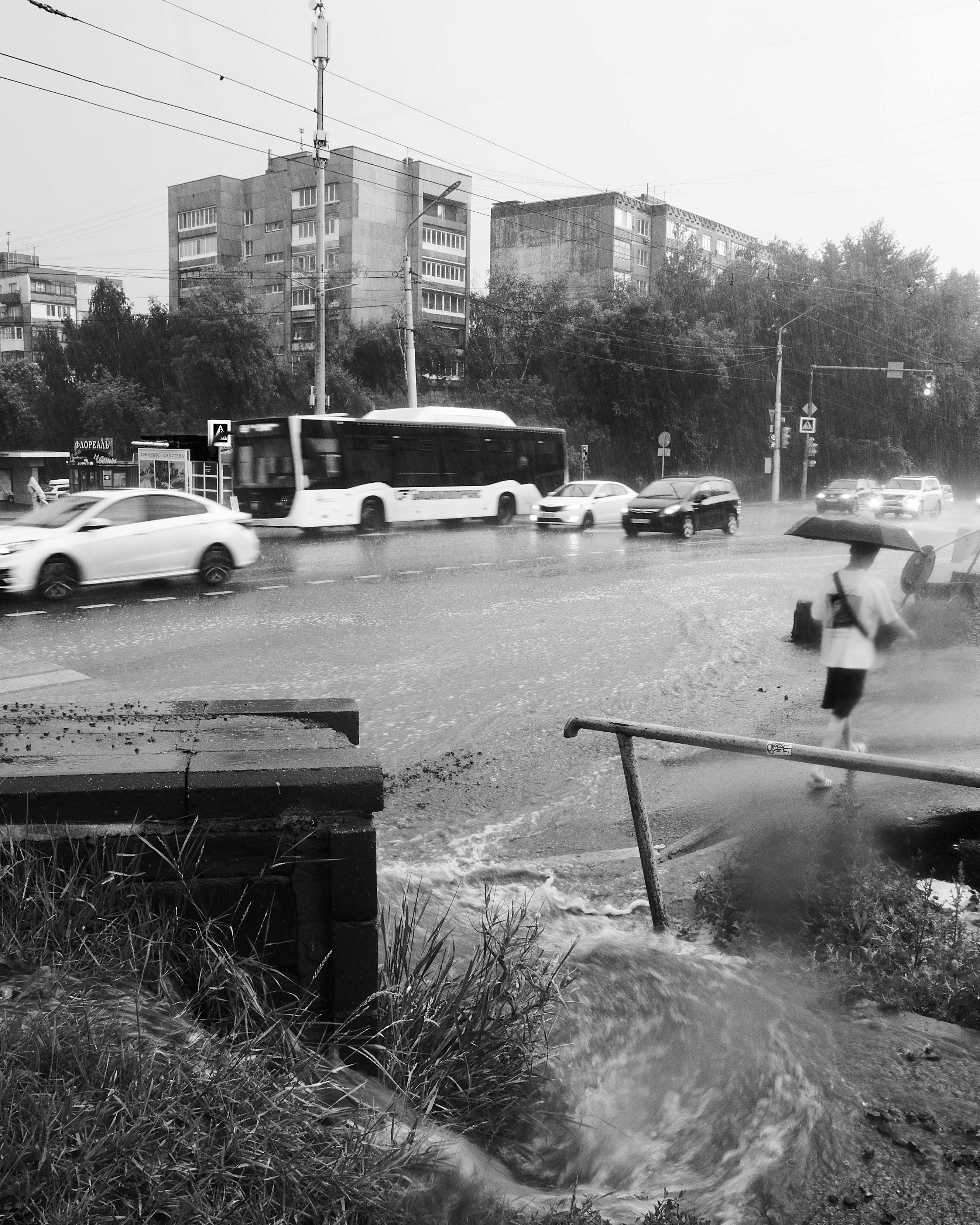 A bustling city intersection drenched in rain, showcasing vehicles splashing through puddles while a pedestrian navigates the downpour with an umbrella.