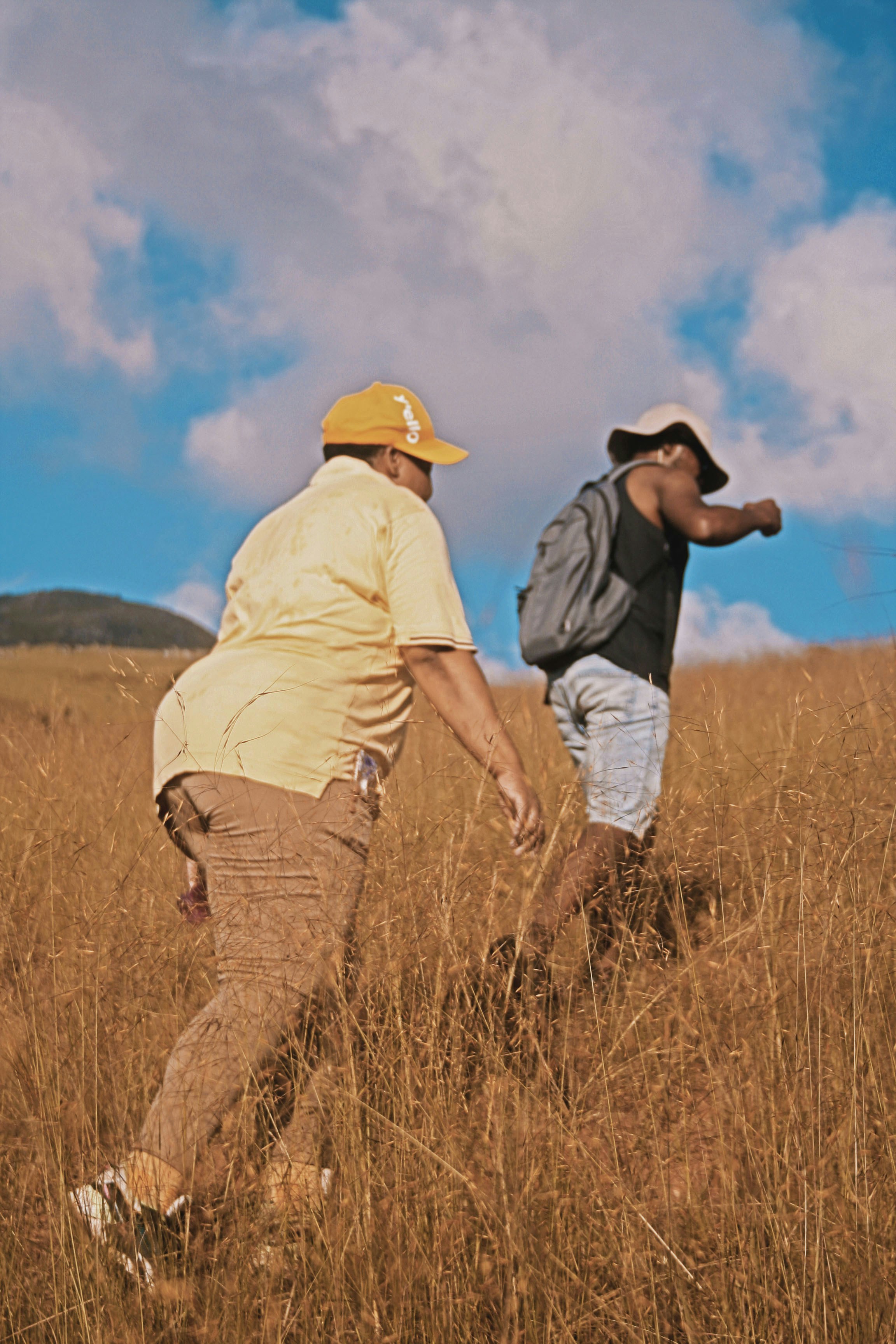 Two hikers ascending a grassy hill under a bright blue sky, with fluffy clouds casting shadows. The scene captures the spirit of adventure and exploration.