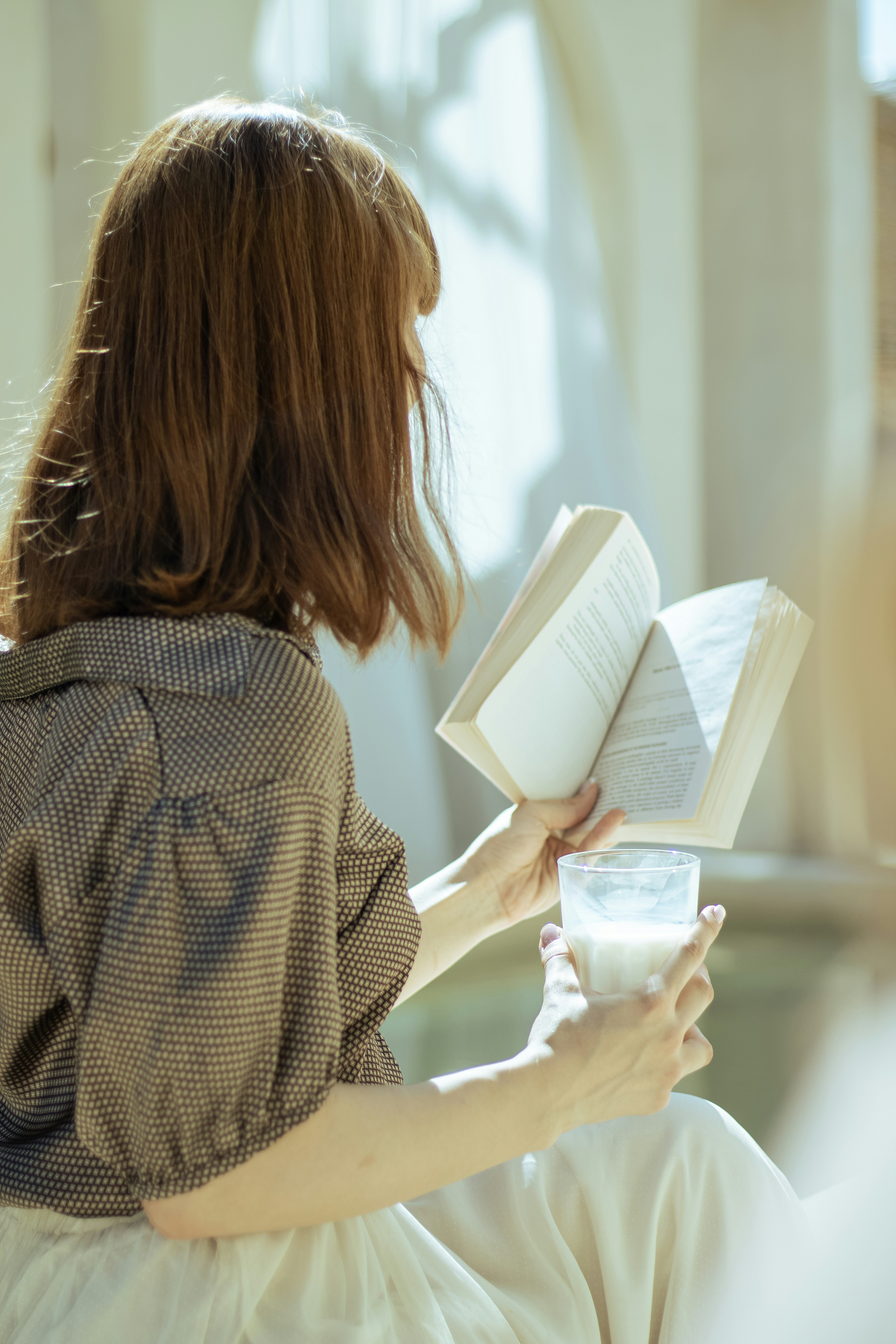 Individual engrossed in a book while holding a glass of milk, seated in a sunlit space with soft shadows.