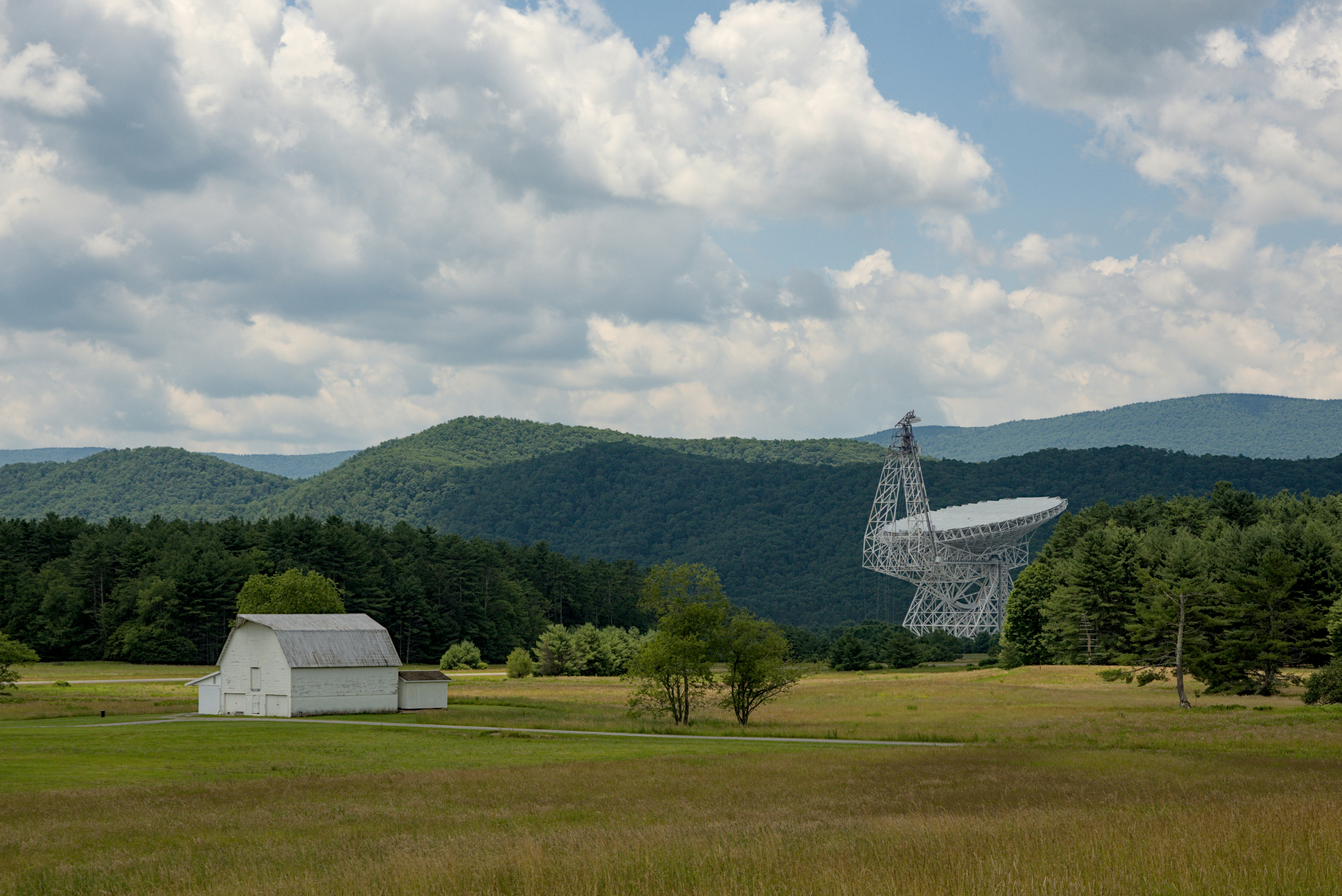 A large radio telescope stands in contrast to a serene countryside scene featuring a white barn and rolling green hills under a cloudy sky.