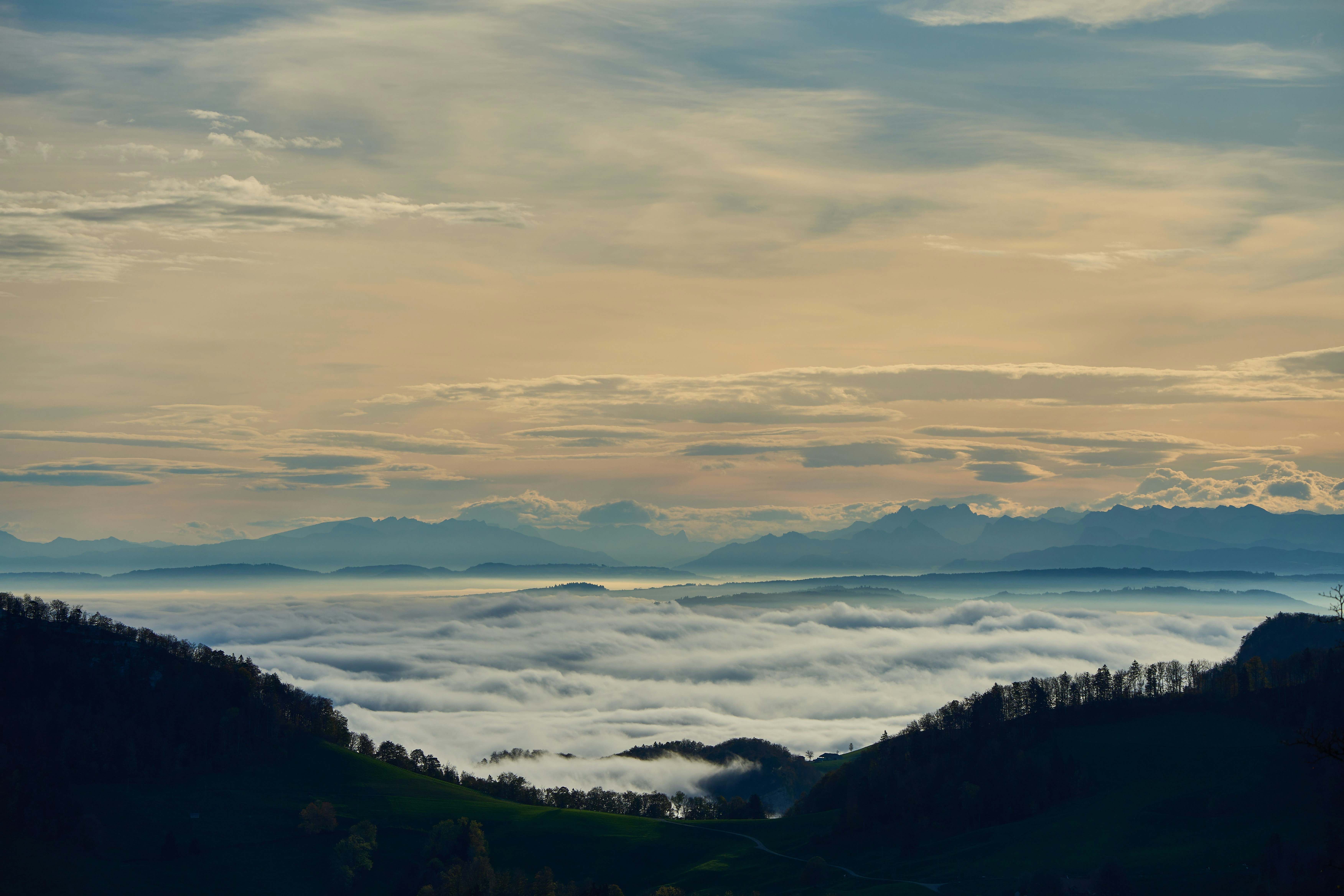 Layered clouds rolling over hills at dusk, with distant mountains faintly visible against a pastel sky.