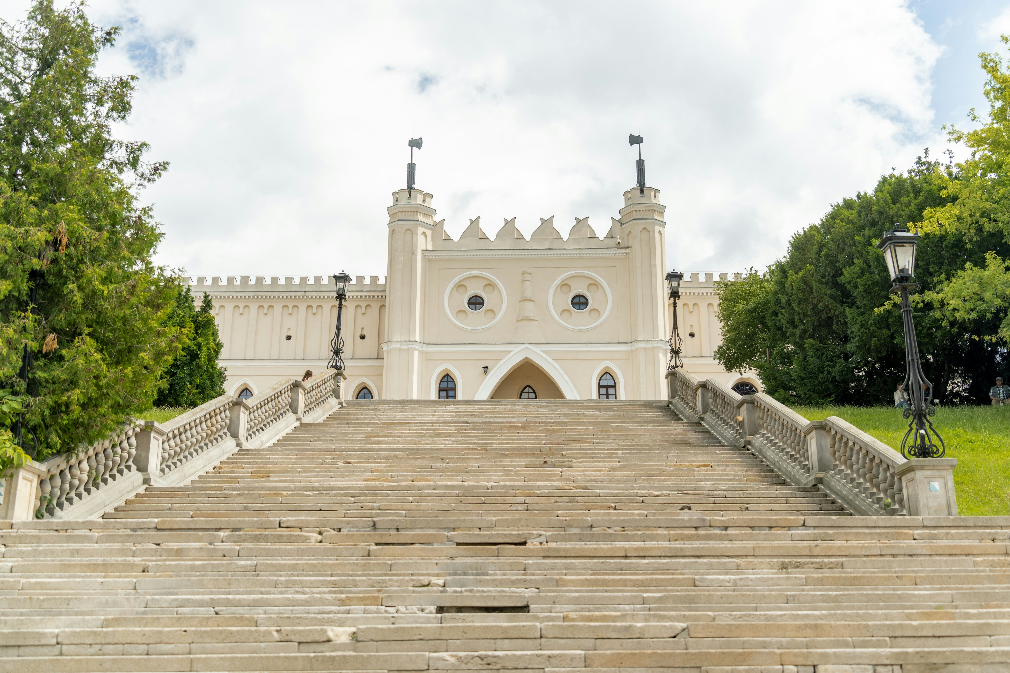 Grand staircase leading to a historic castle-like structure, framed by lush greenery and ornate street lamps.