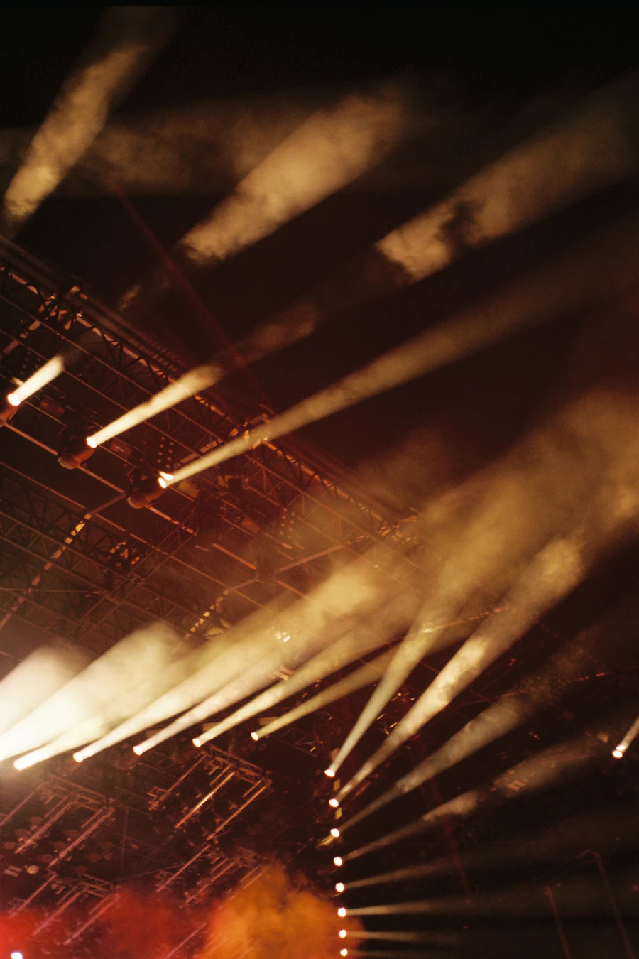 Warm beams of stage lighting cut through heavy haze beneath a metal truss ceiling, creating a dramatic abstract concert scene.