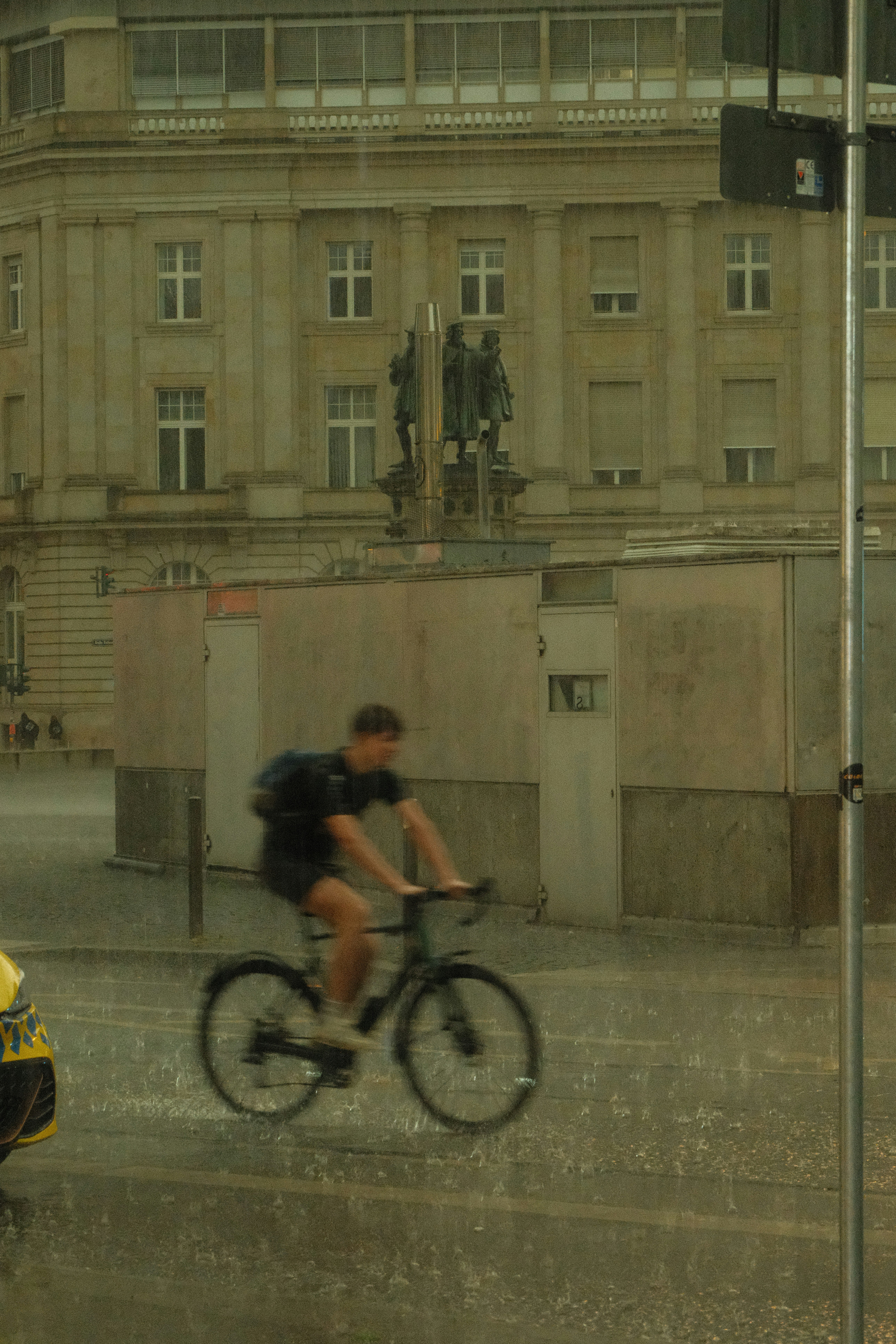 A cyclist navigates through heavy rain in a bustling urban environment, with a statue and historic architecture in the background.