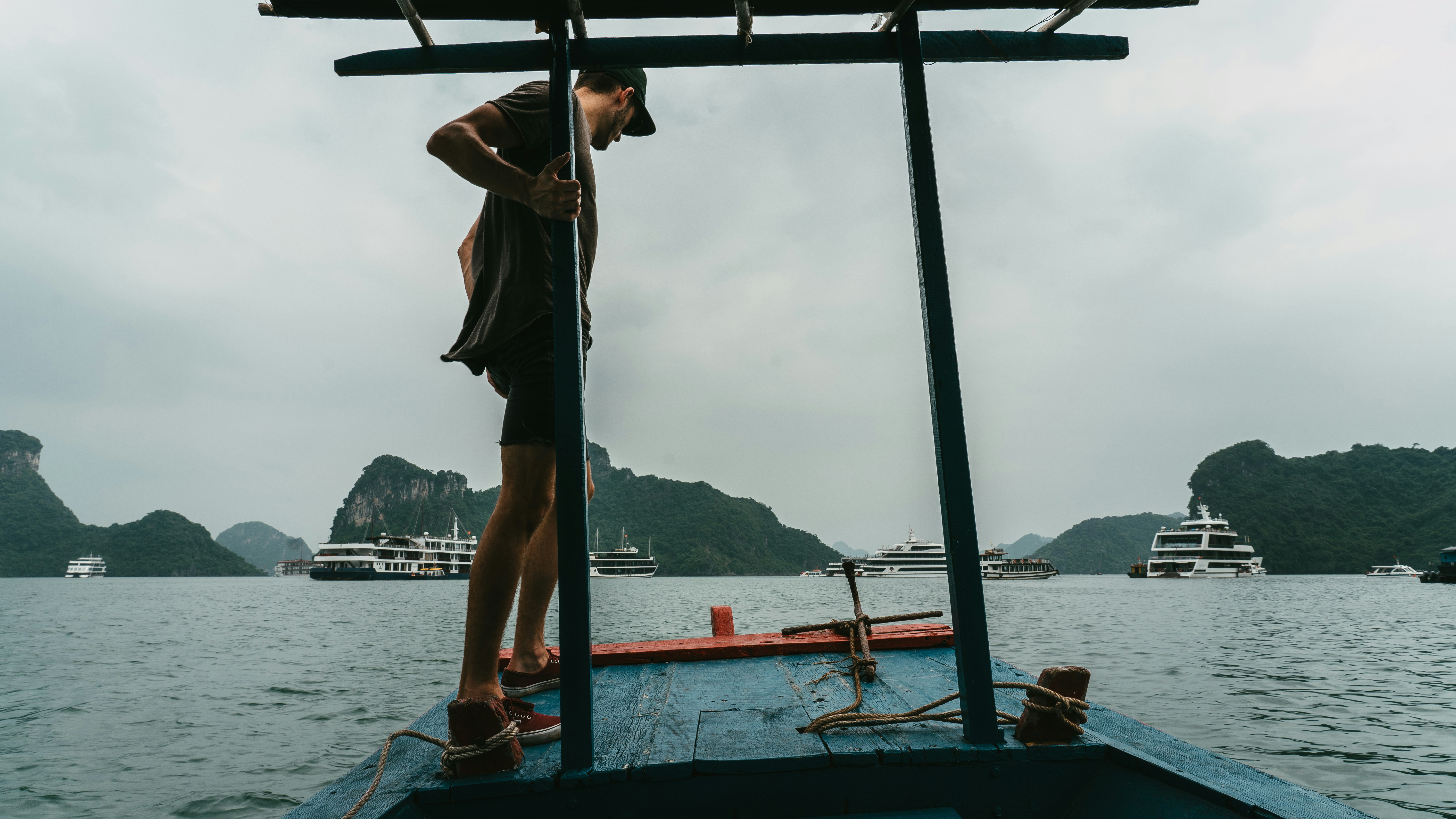 A person stands on a wooden boat, gazing at the serene landscape of limestone islands and distant boats in a misty bay.