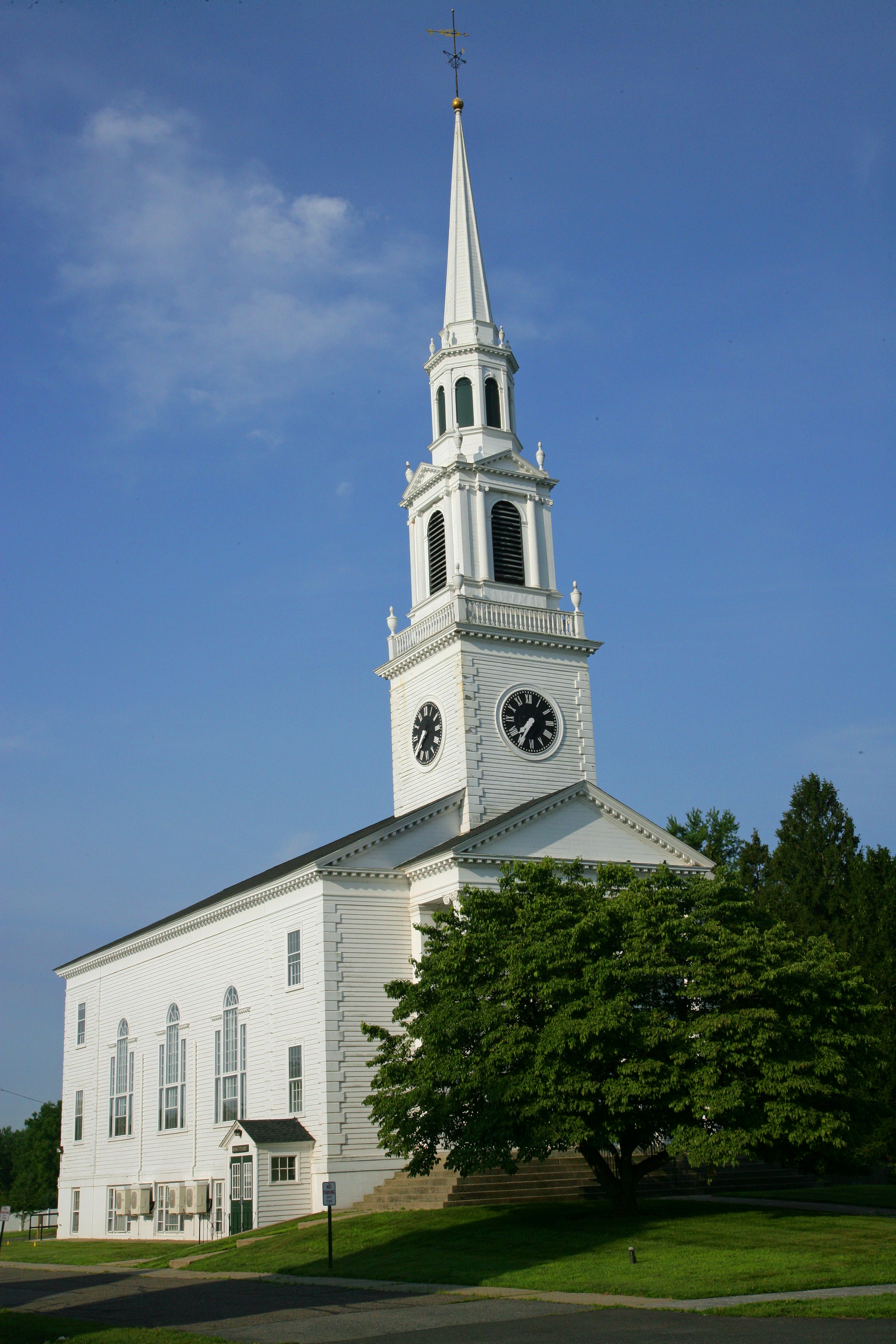 Historic white church with a tall steeple and large clock, surrounded by lush greenery under a clear blue sky.