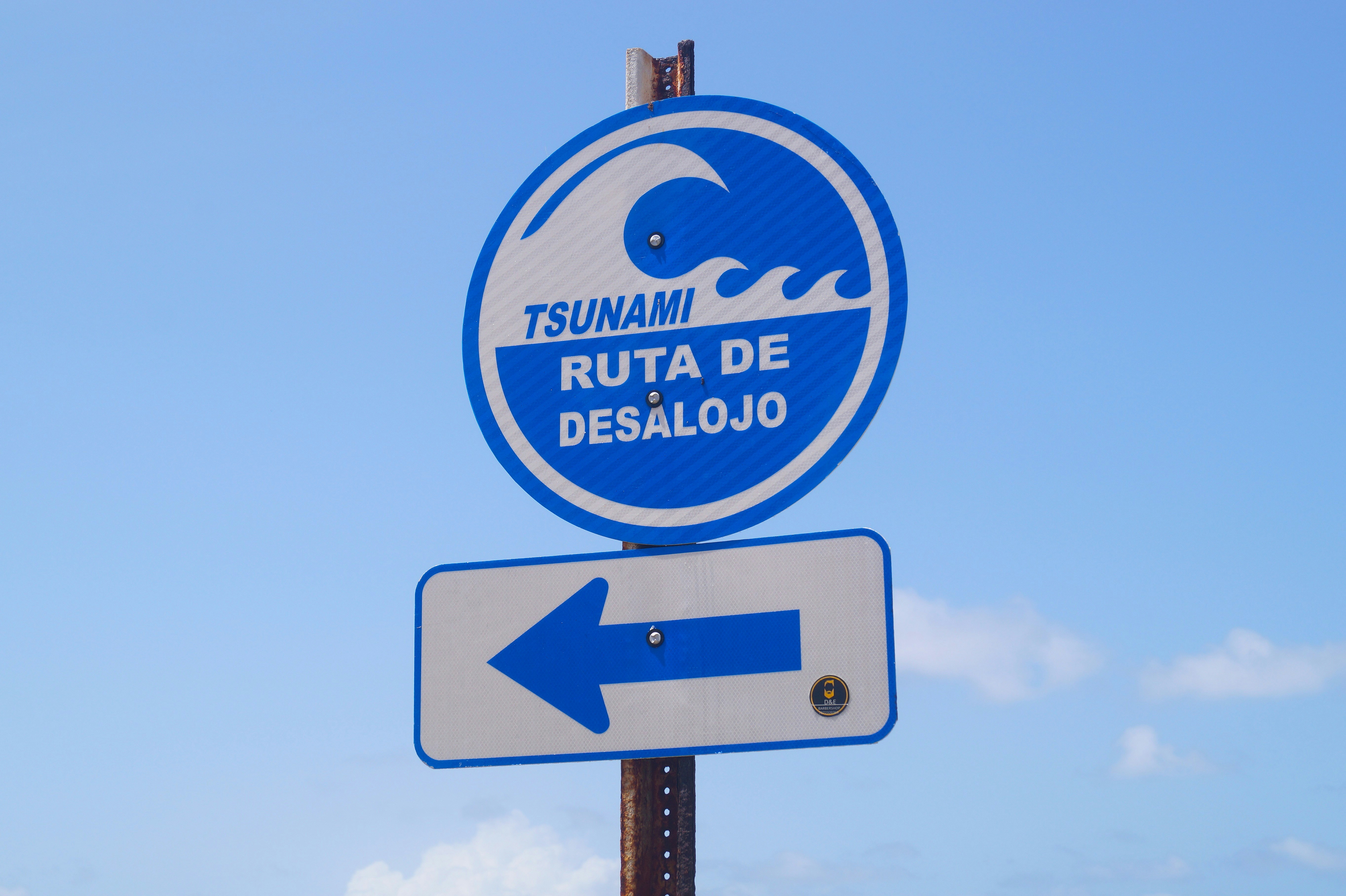 Spanish Tsunami Sign at the beach with blue sky in the background. | Tsunami evacuation route sign pointing left.
