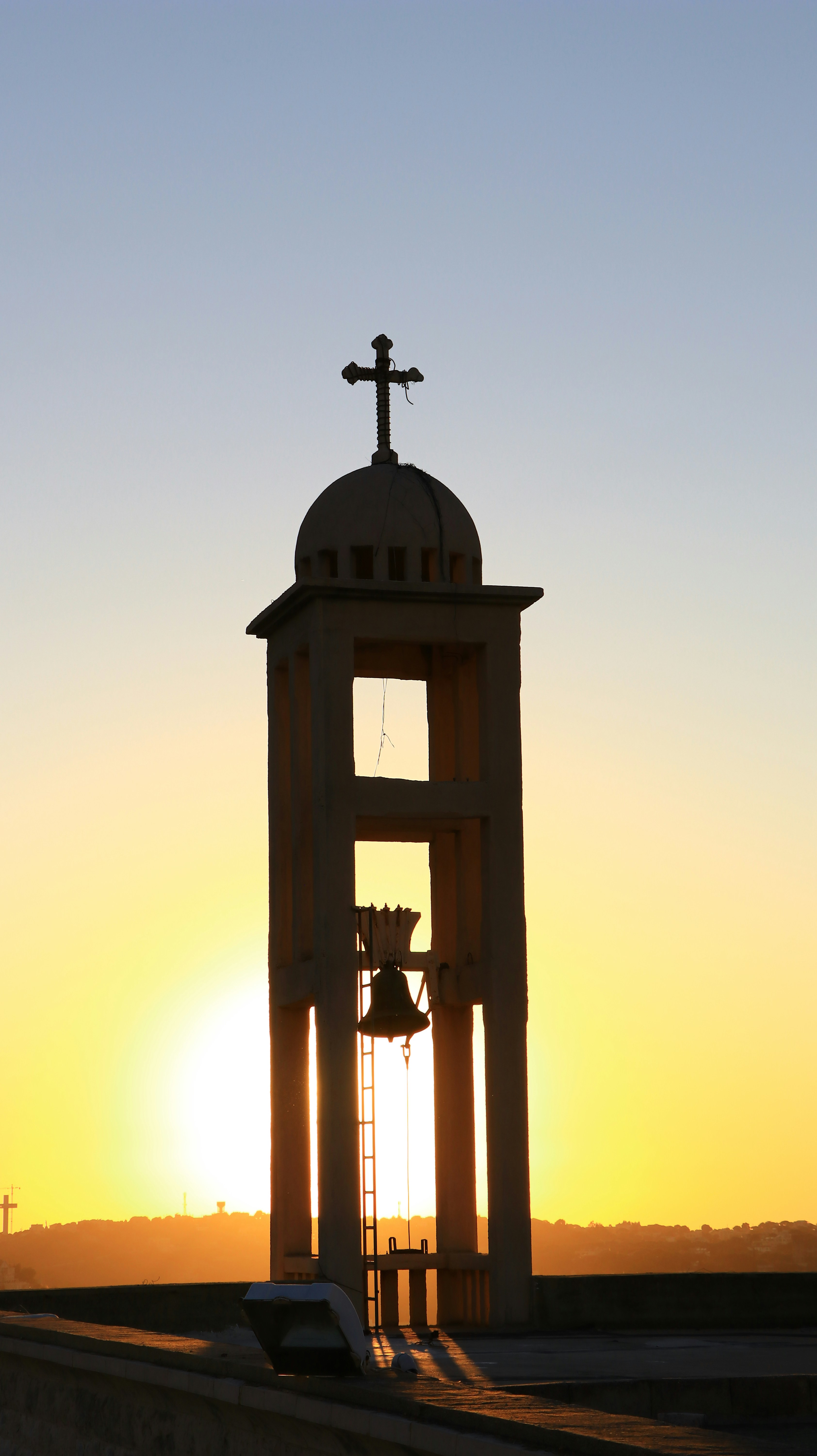 Silhouette of a bell tower against a vibrant sunrise, featuring a cross atop and a bell hanging inside. The scene captures the serene transition from night to day.