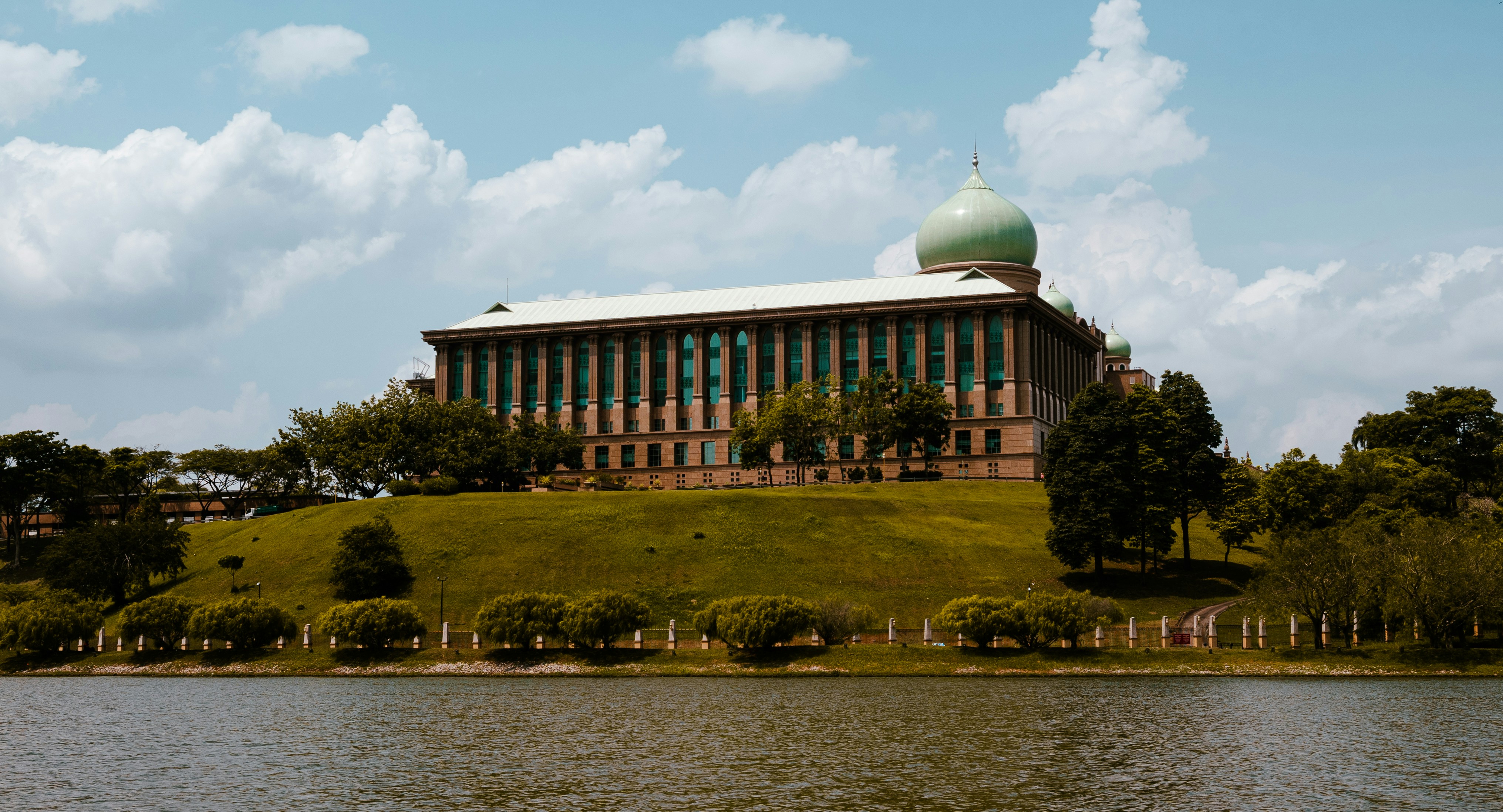 A grand building with a green dome stands majestically on a hill, surrounded by lush greenery and reflecting in the tranquil waters below.