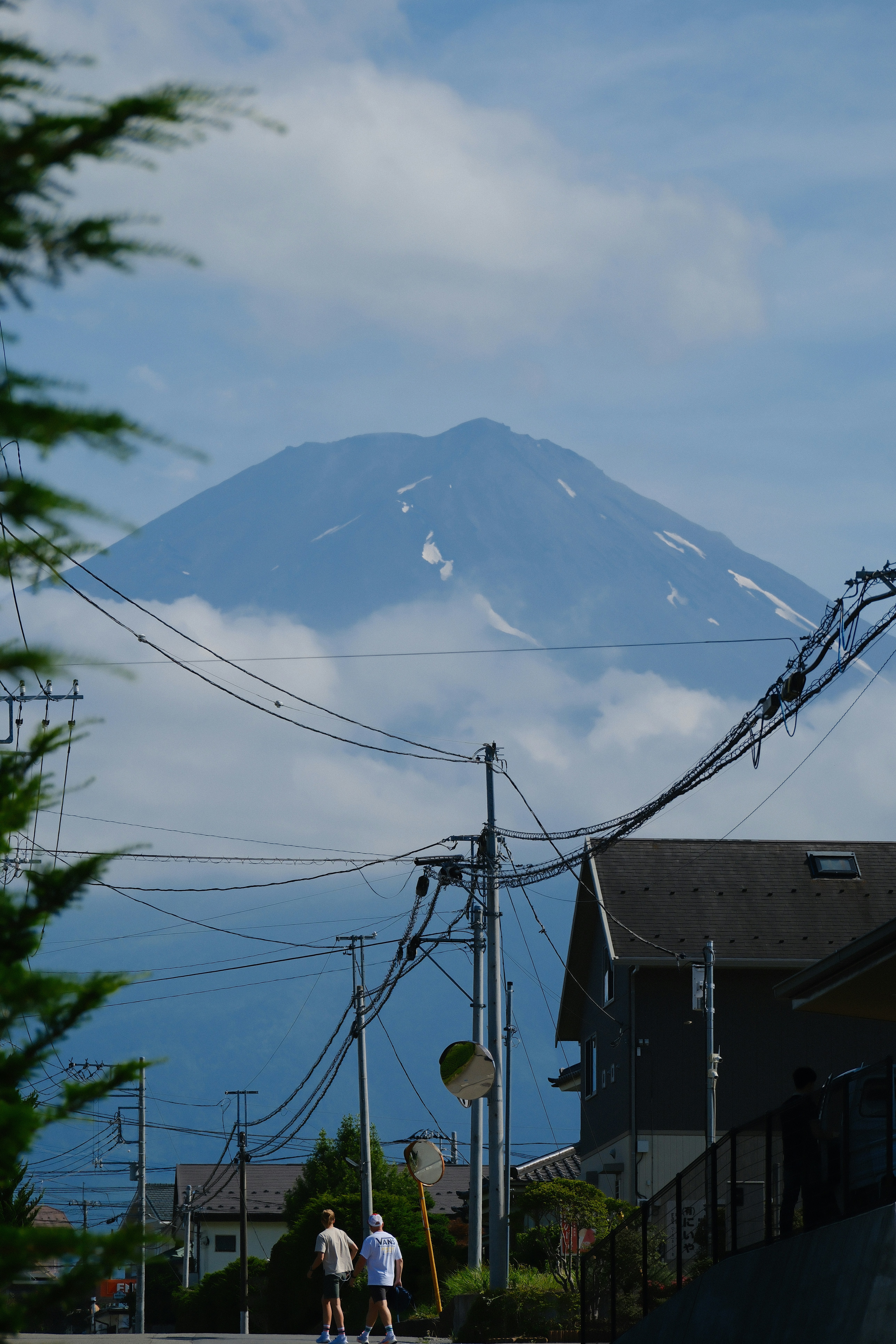 Two individuals walking along a street with a prominent mountain looming in the background, framed by power lines and greenery.