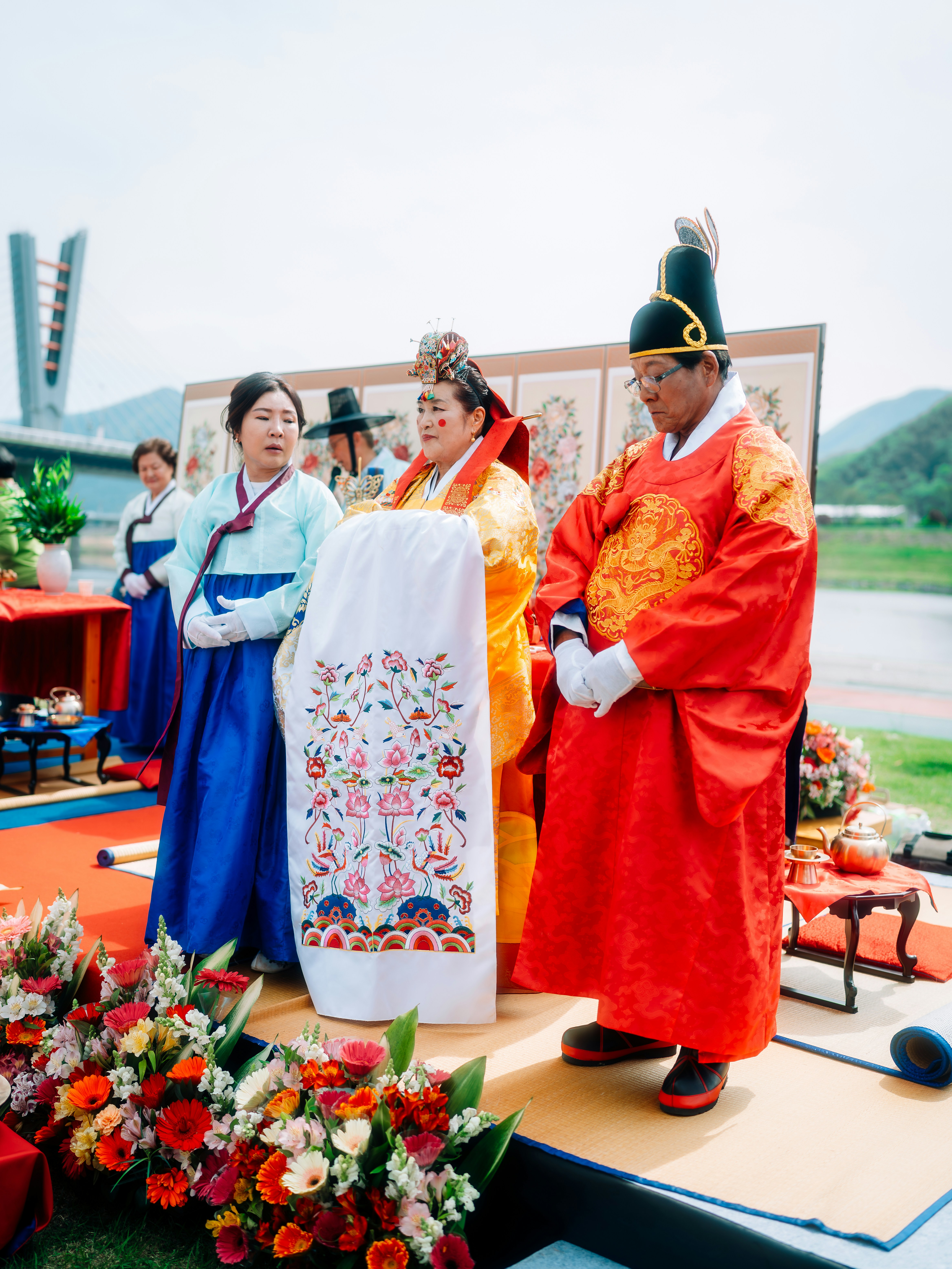 Participants in traditional attire engage in a ceremonial event, surrounded by vibrant floral arrangements and a scenic backdrop. The focus is on the rich cultural heritage displayed through clothing and rituals.