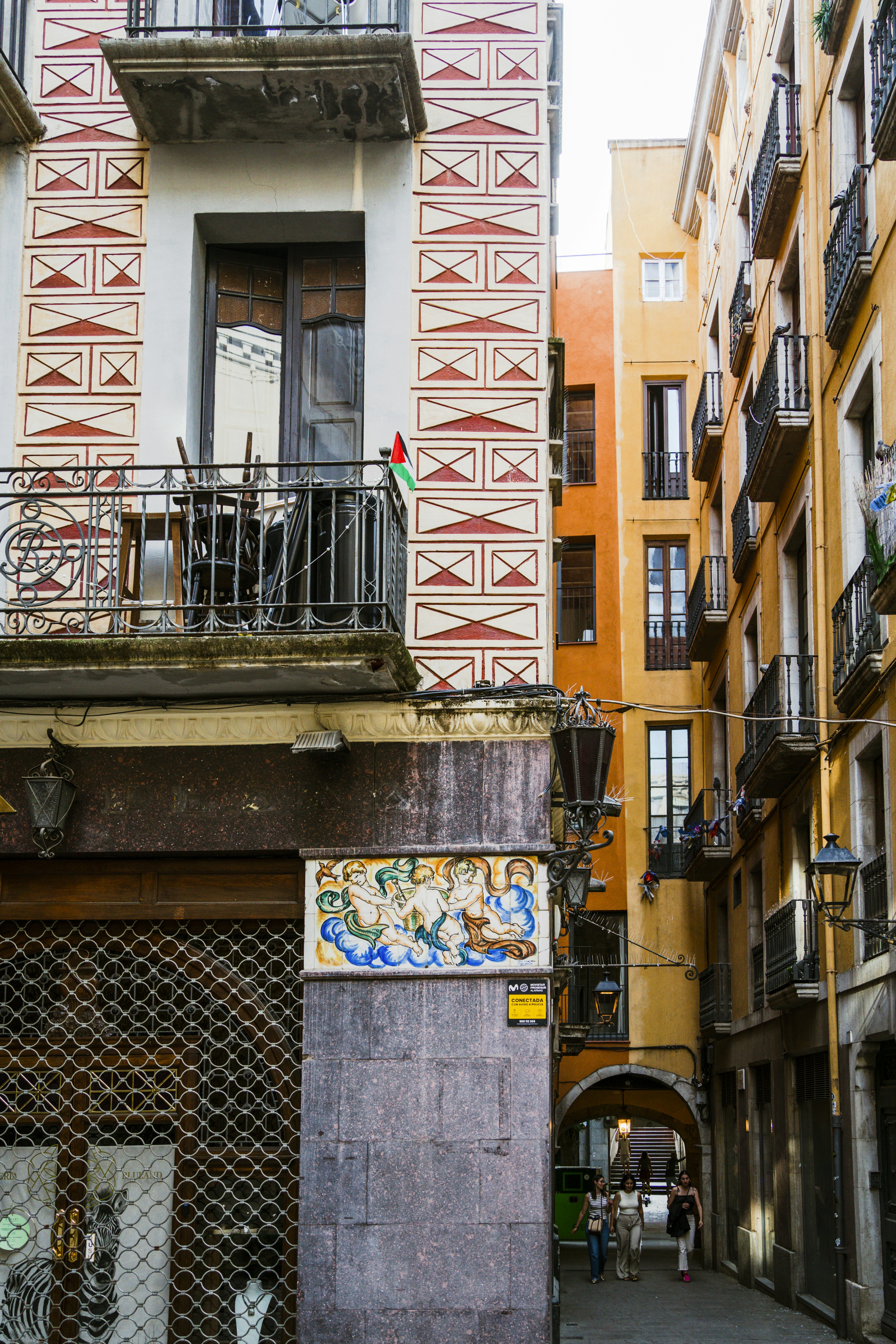 Intricate balcony overlooking a vibrant alleyway, showcasing architectural details and a glimpse of daily life in a bustling city. The ornate mural adds a touch of artistry to the scene.