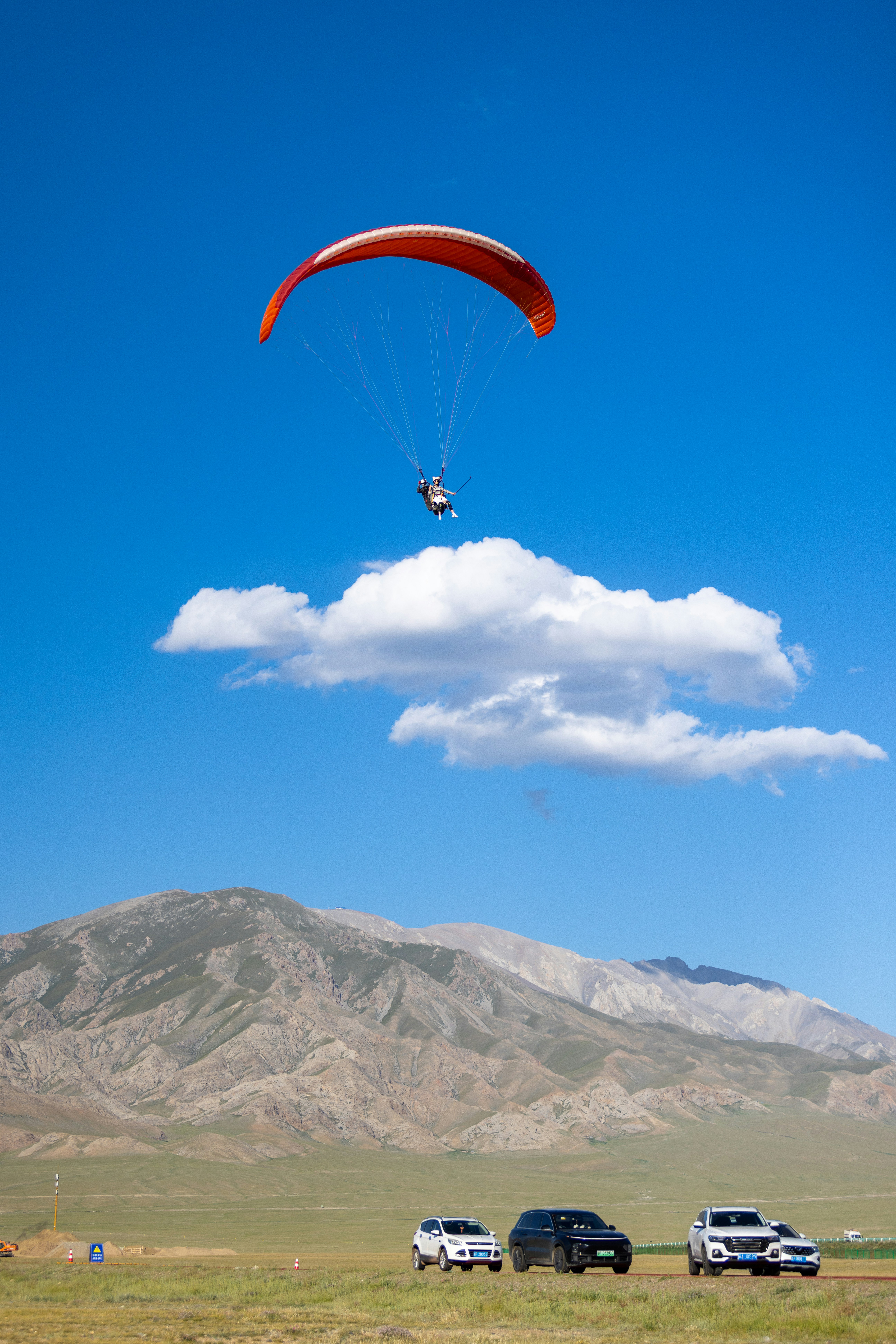 A paraglider gracefully floats in the sky, with a vibrant orange canopy against a backdrop of fluffy clouds and majestic mountains. Three vehicles are parked below, adding scale to the scene.