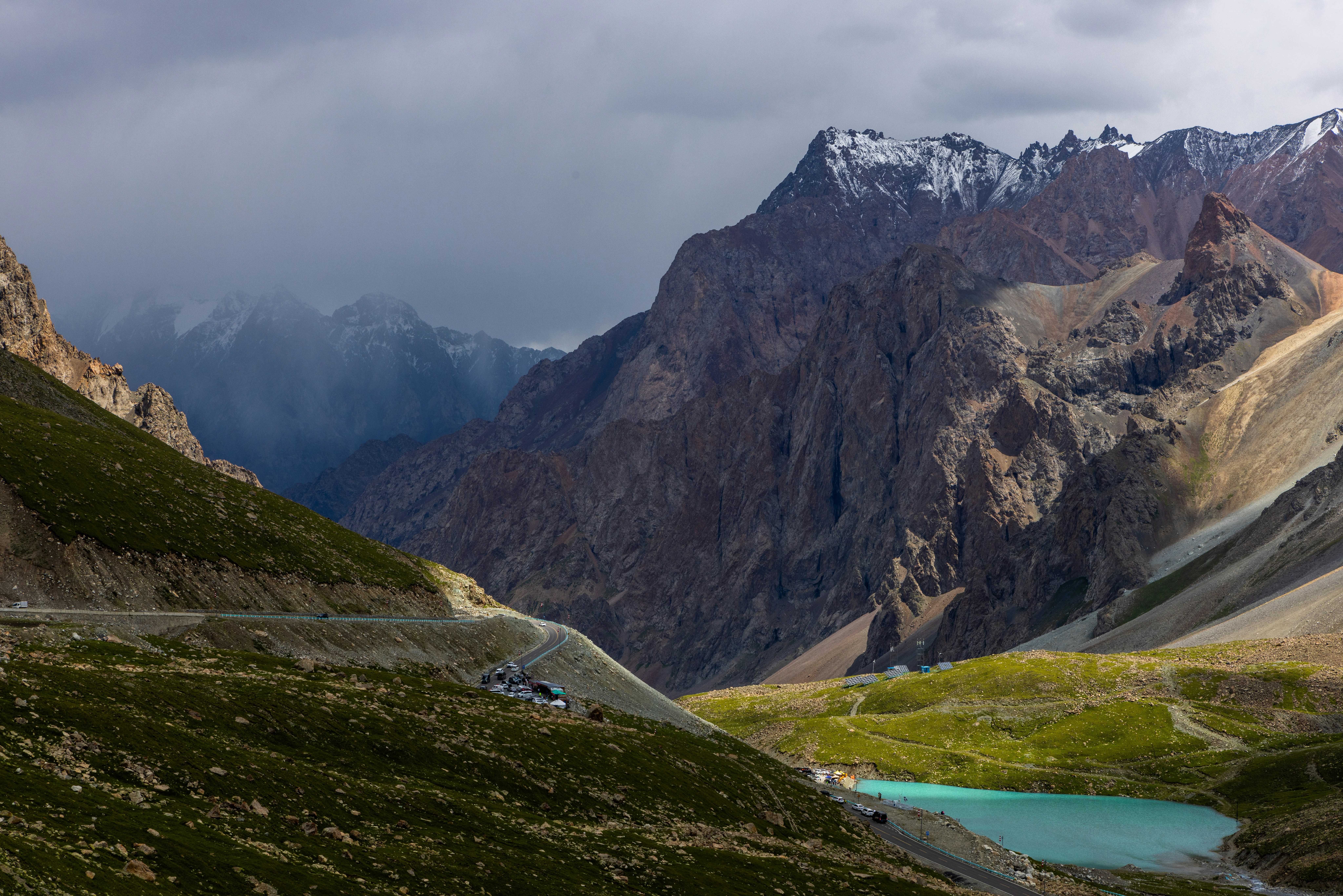 Dramatic mountain range under a moody sky, with a winding road leading to a serene turquoise lake nestled in the valley. The rugged terrain showcases nature's raw beauty.
