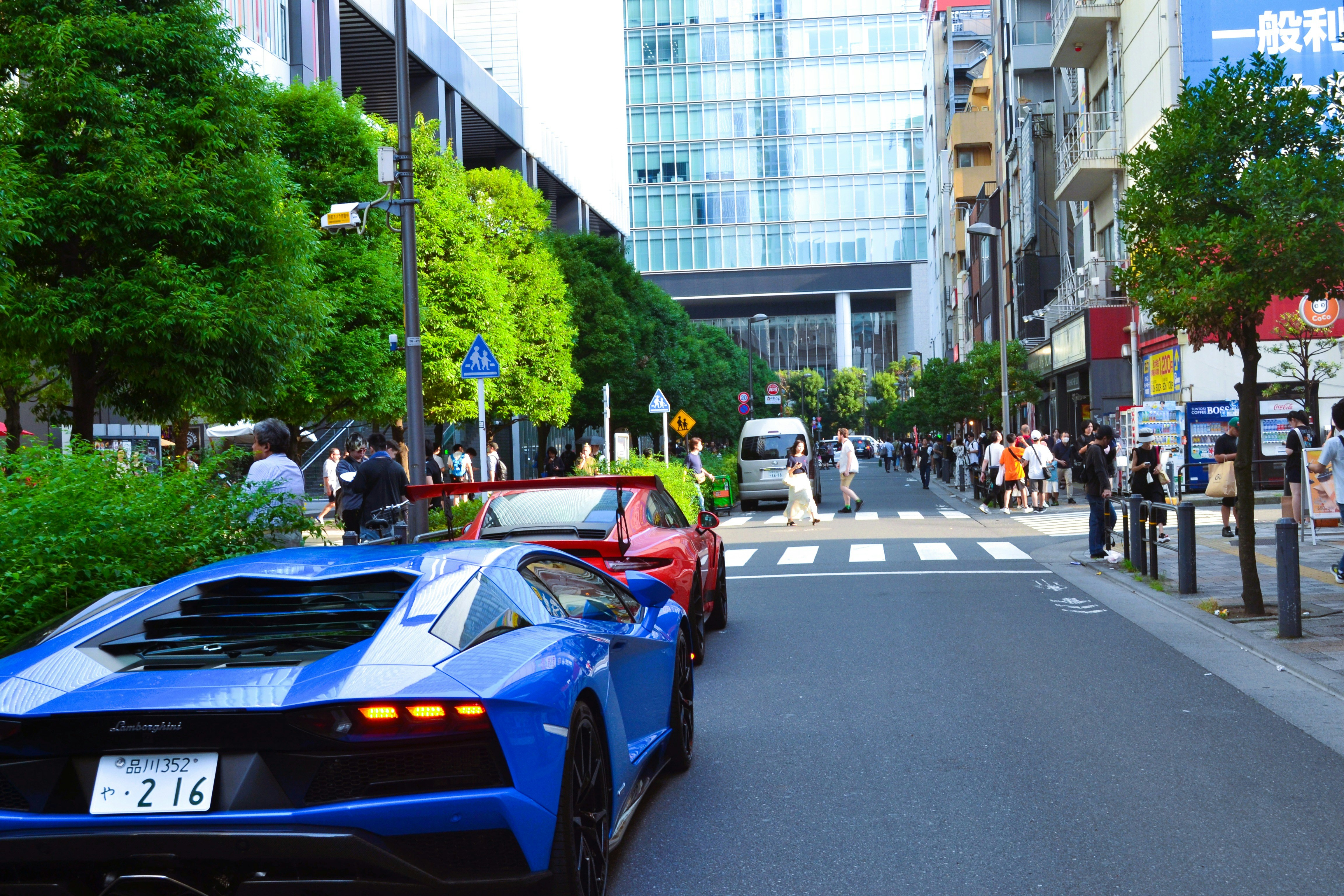 Two luxurious sports cars parked on a bustling city street lined with greenery and pedestrians. The scene captures the vibrant energy of urban life.