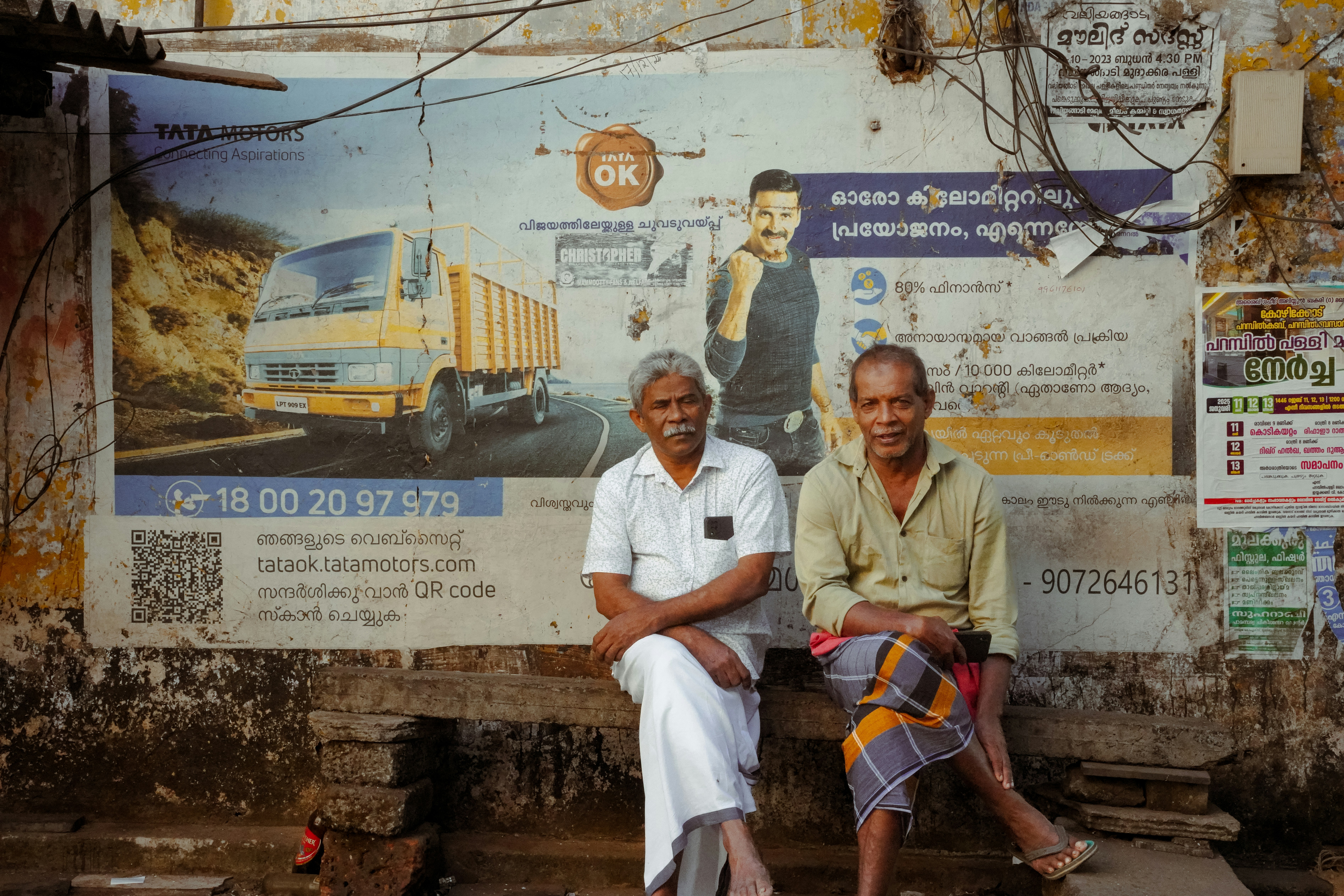 Simple people from Kerala | Two men sit in front of a weathered advertisement.
