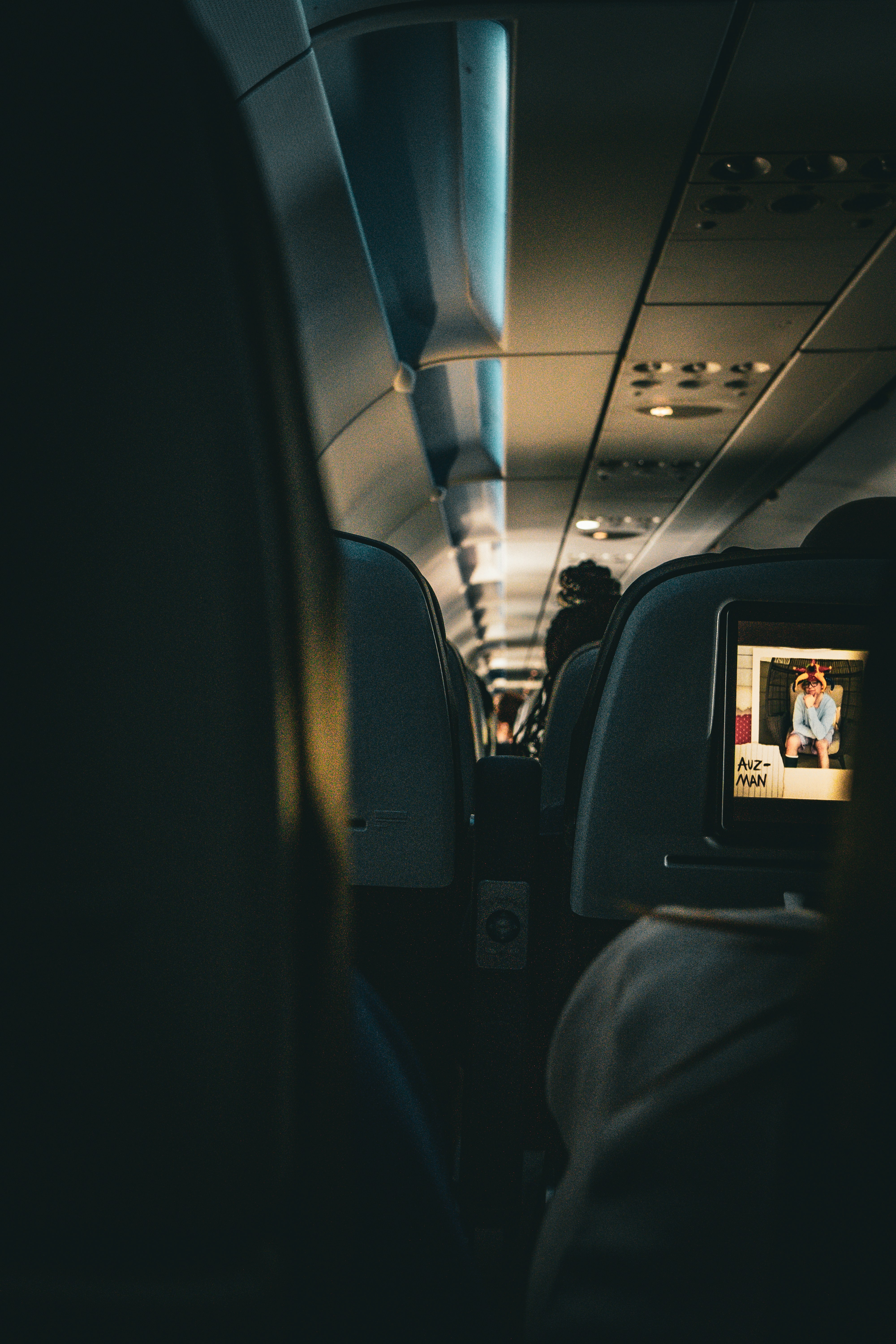 Passengers in an airplane cabin, with one viewing content on a personal screen. The atmosphere is dimly lit, emphasizing the intimate setting.