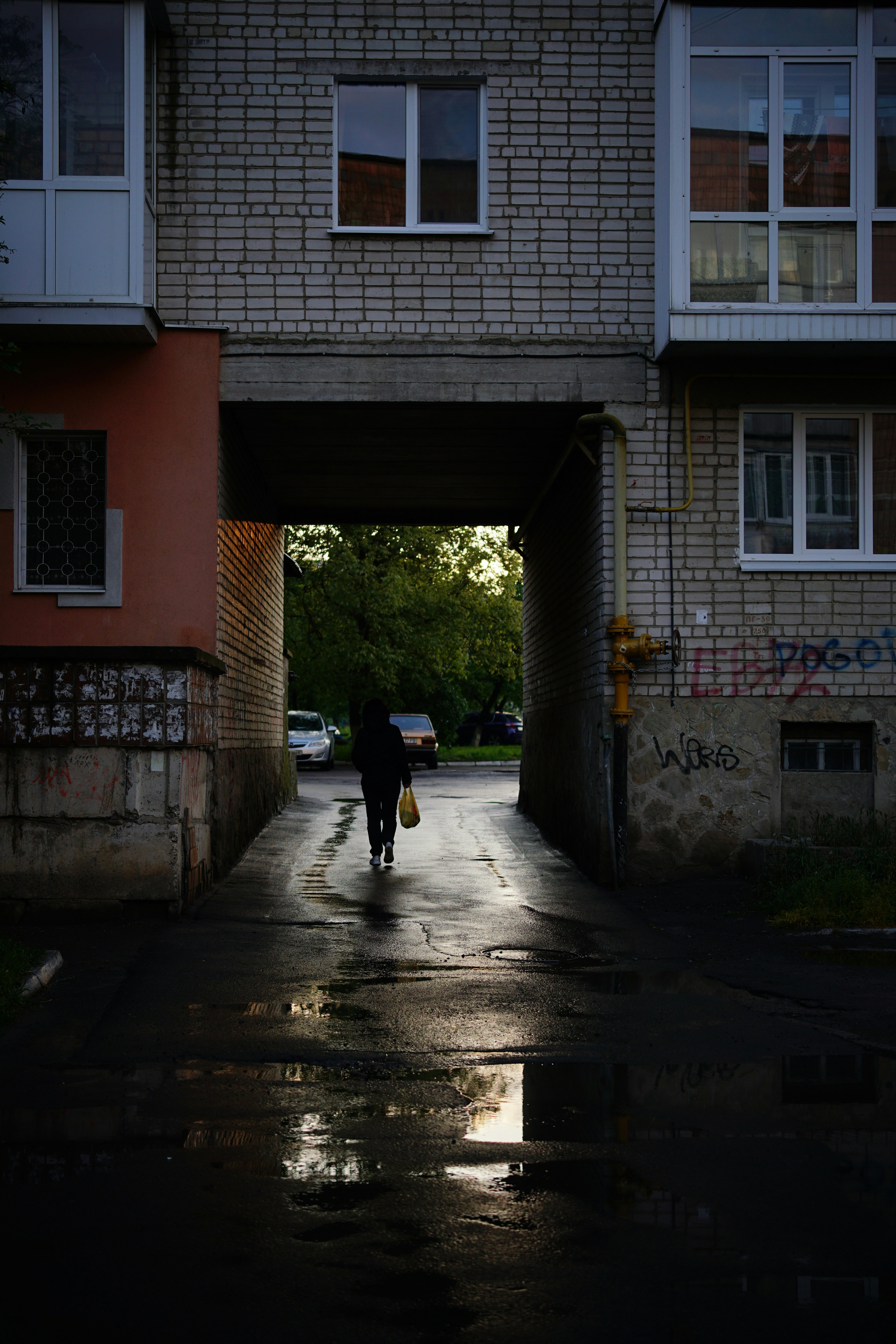 Silhouette of a person walking under an archway with reflections on wet pavement, framed by urban architecture. The scene evokes a sense of quiet contemplation.