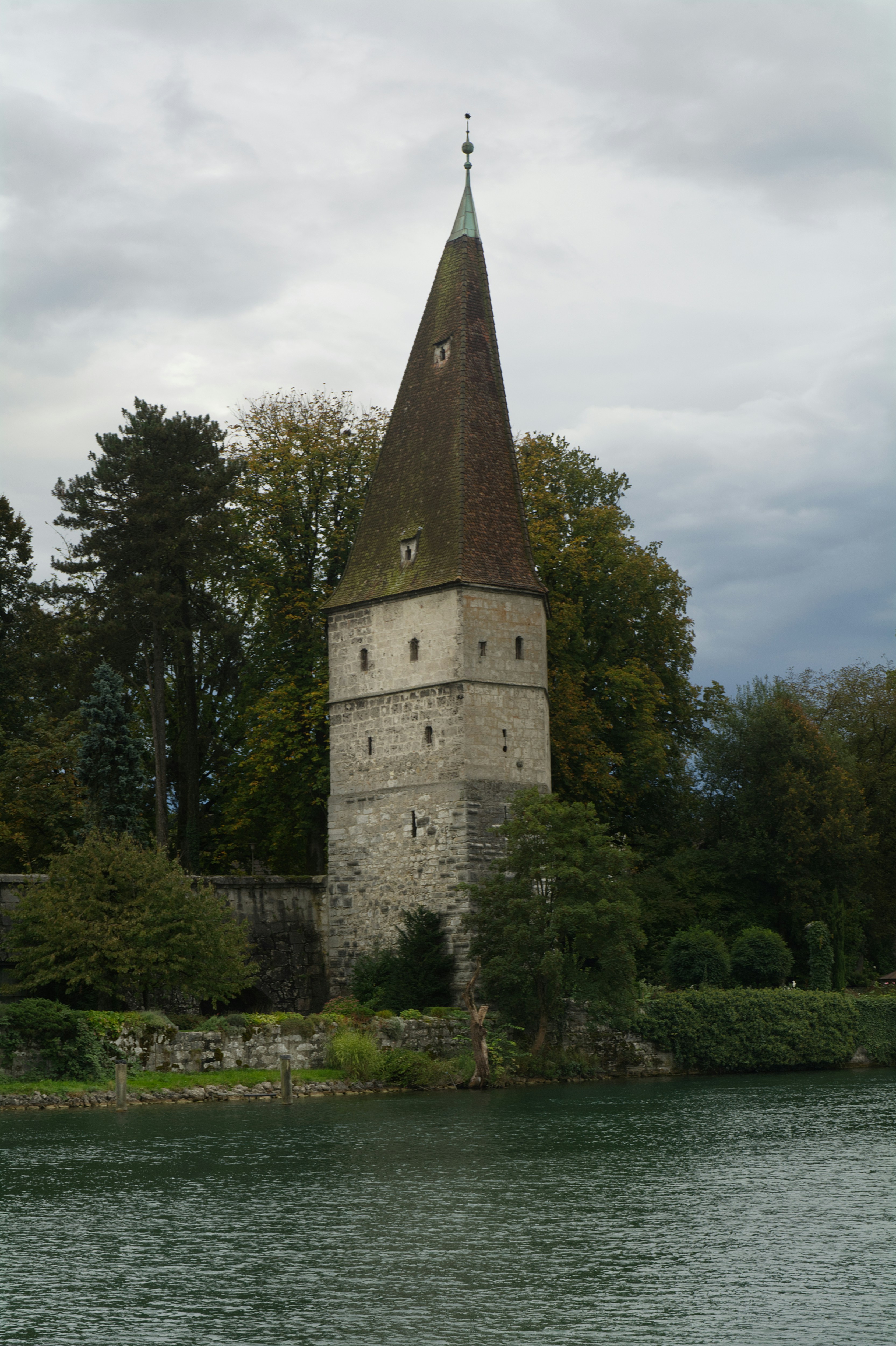 Crooked tower "Krummturm" seen from across river Aare | A stone tower stands beside the water.