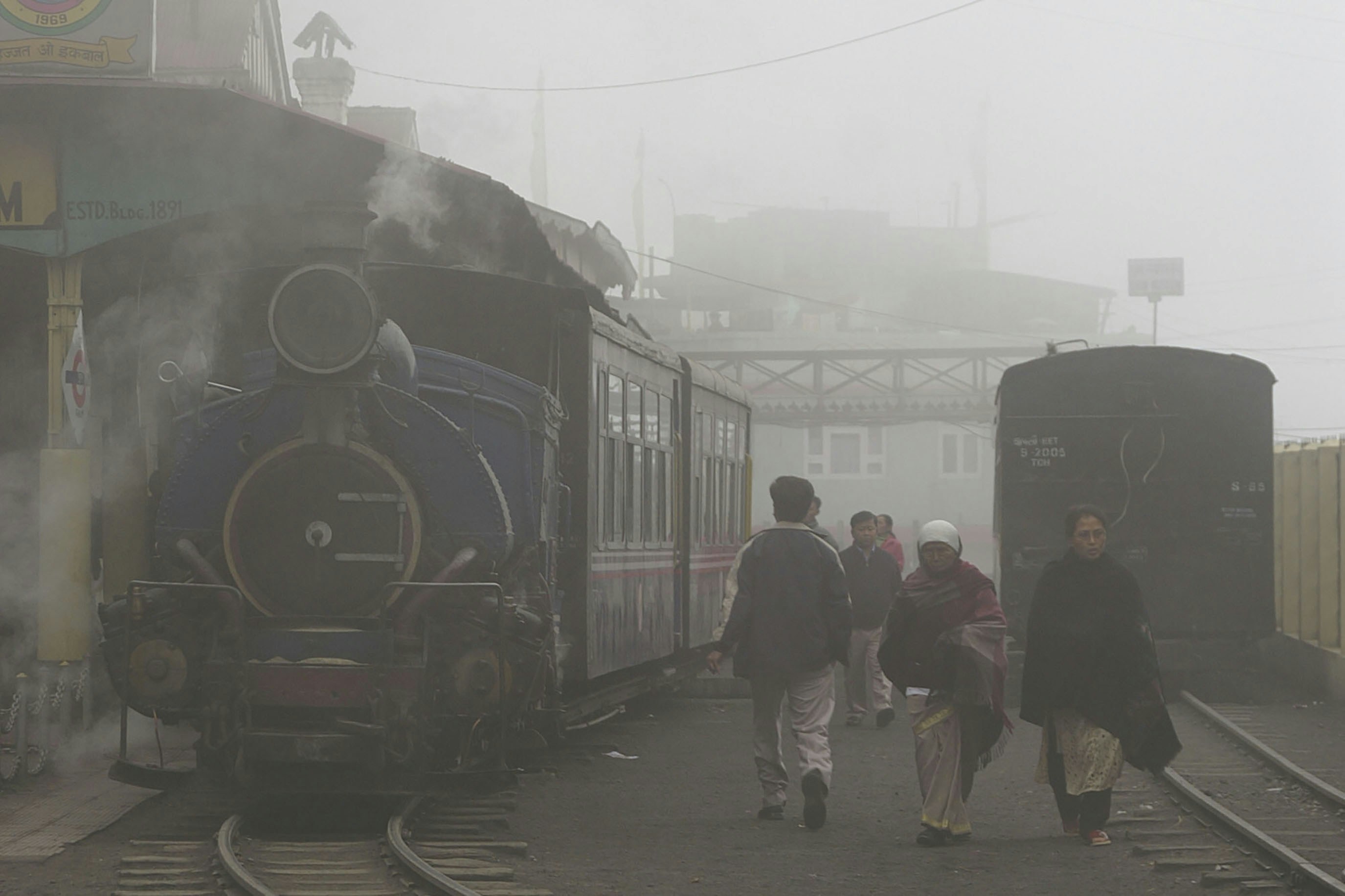Steam locomotive surrounded by pedestrians in a foggy railway station, evoking a sense of nostalgia and adventure.