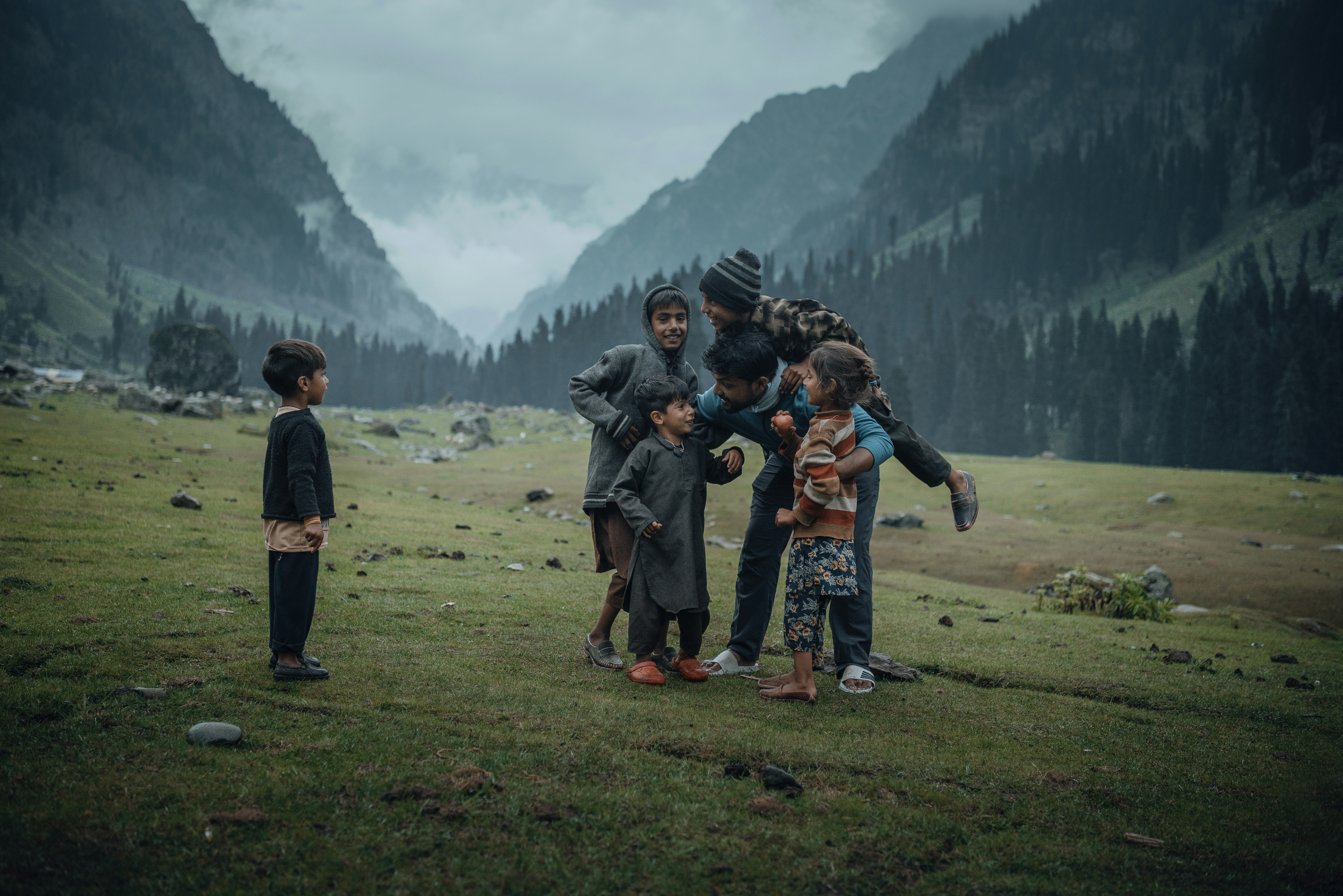 Group of children playing together in a lush green valley surrounded by mountains, with one child being lifted joyfully by another. 