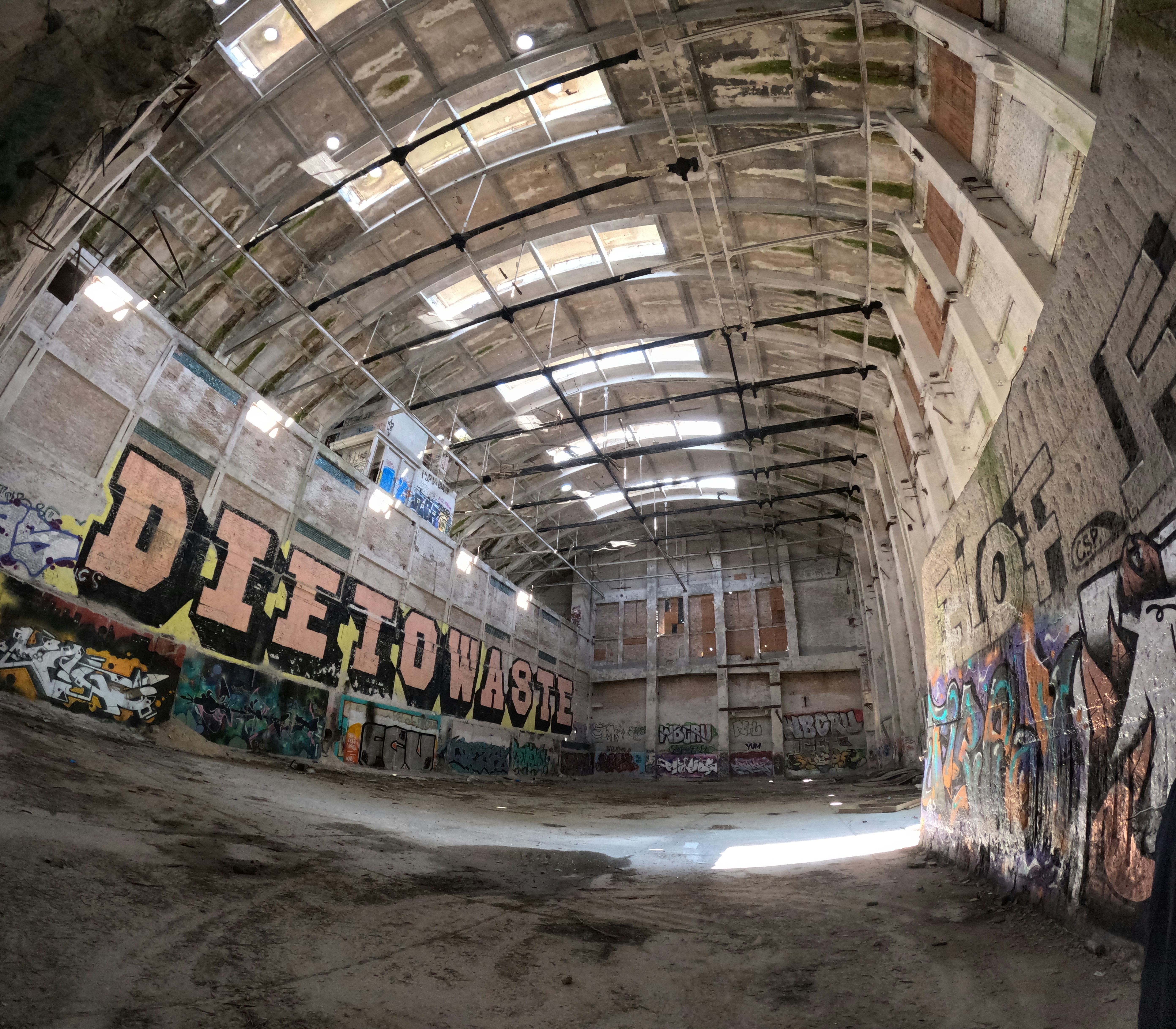 Interior of an abandoned warehouse adorned with vibrant graffiti, showcasing the interplay of light and shadow. The large mural reads 'DIETOWASTE'.