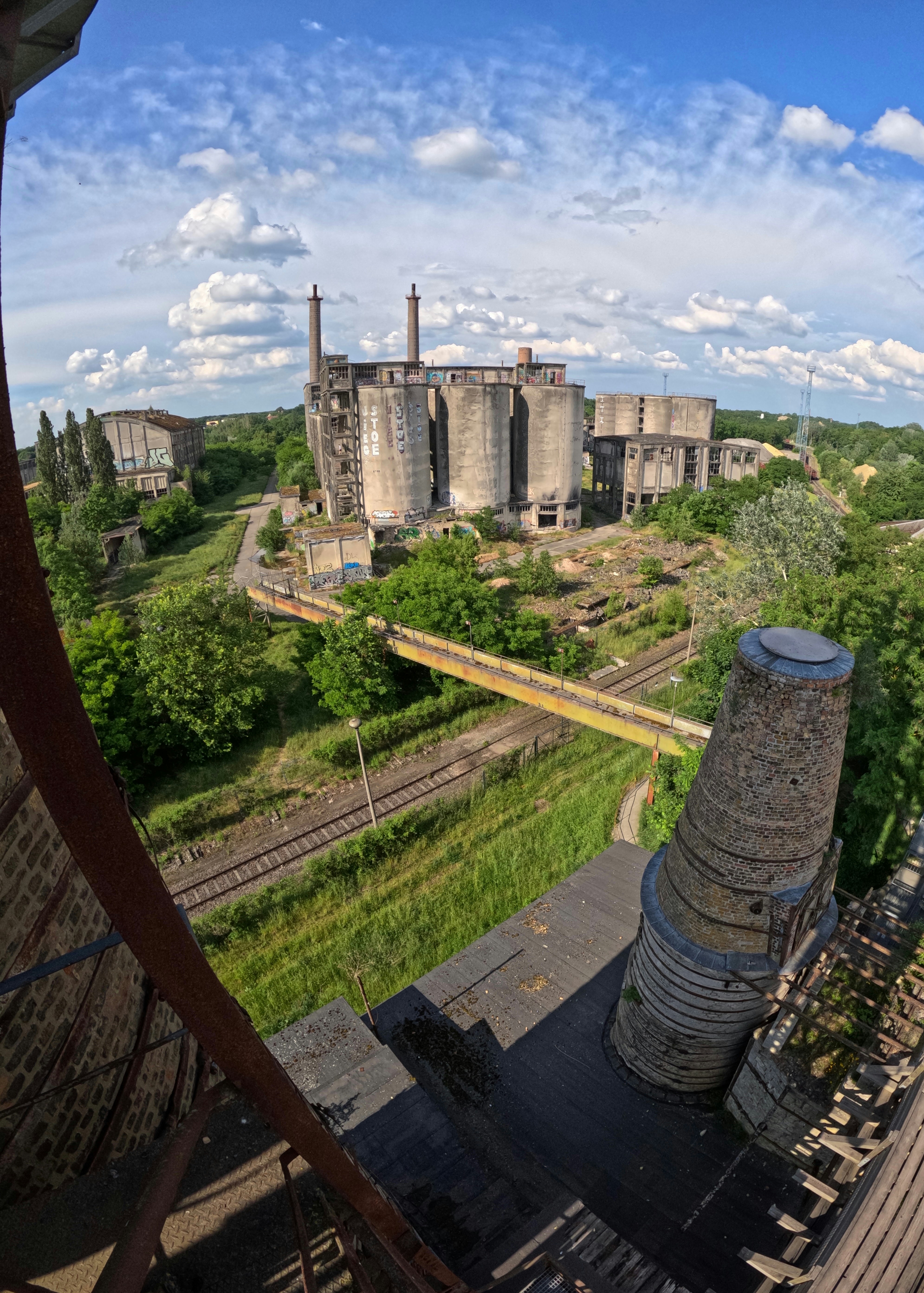 Abandoned industrial complex surrounded by lush greenery and remnants of rail tracks. The scene captures the contrast between nature and human-made structures.