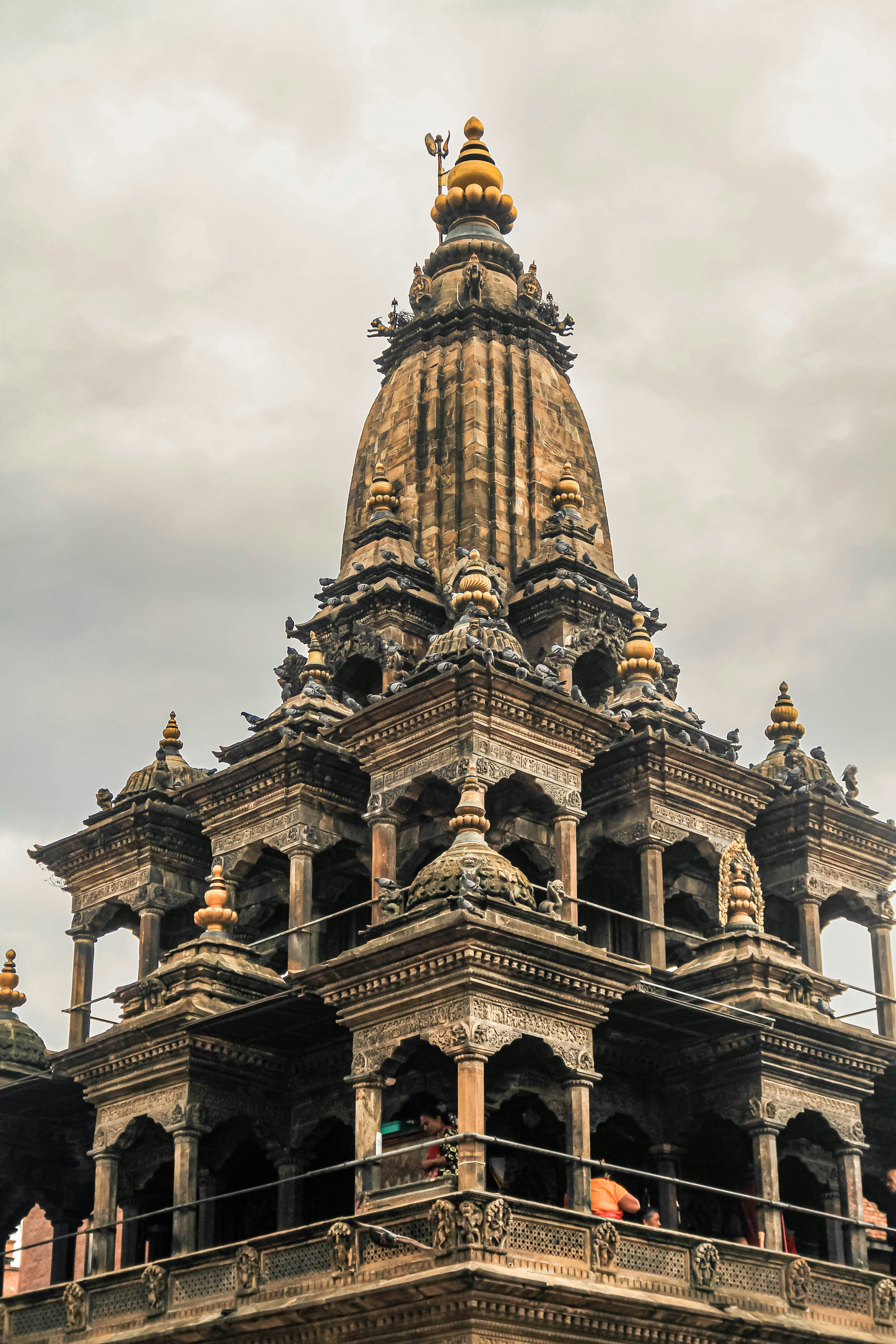A close-up view of the majestic Krishna Mandir in Patan Durbar Square, Lalitpur, Nepal. | An ornate temple rises towards a cloudy sky.