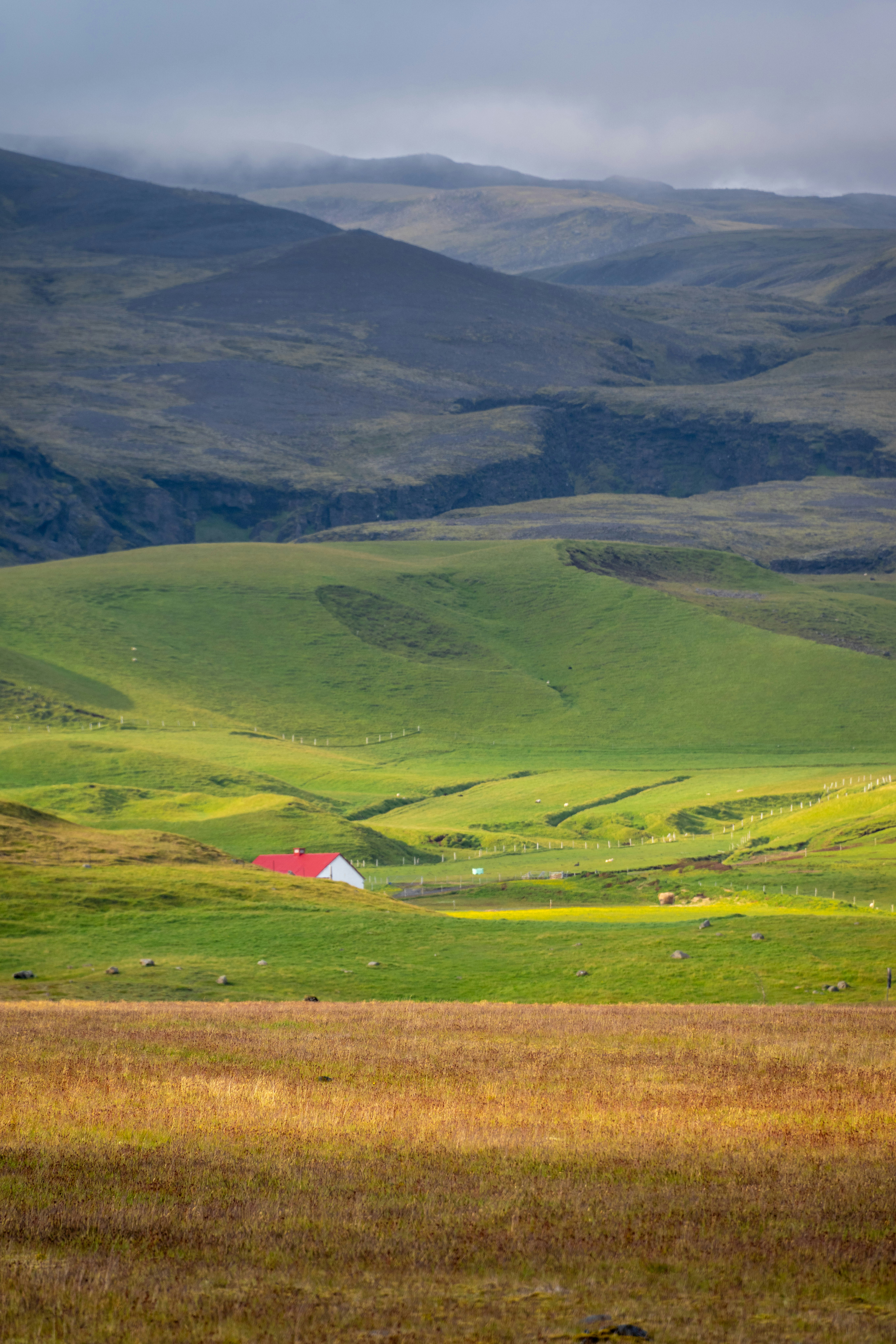 A solitary white farmhouse with a vibrant red roof nestles among rolling emerald hills beneath misty mountains at Dyrhólaey on Iceland’s southern coast. | A house with a red roof is in a green valley.