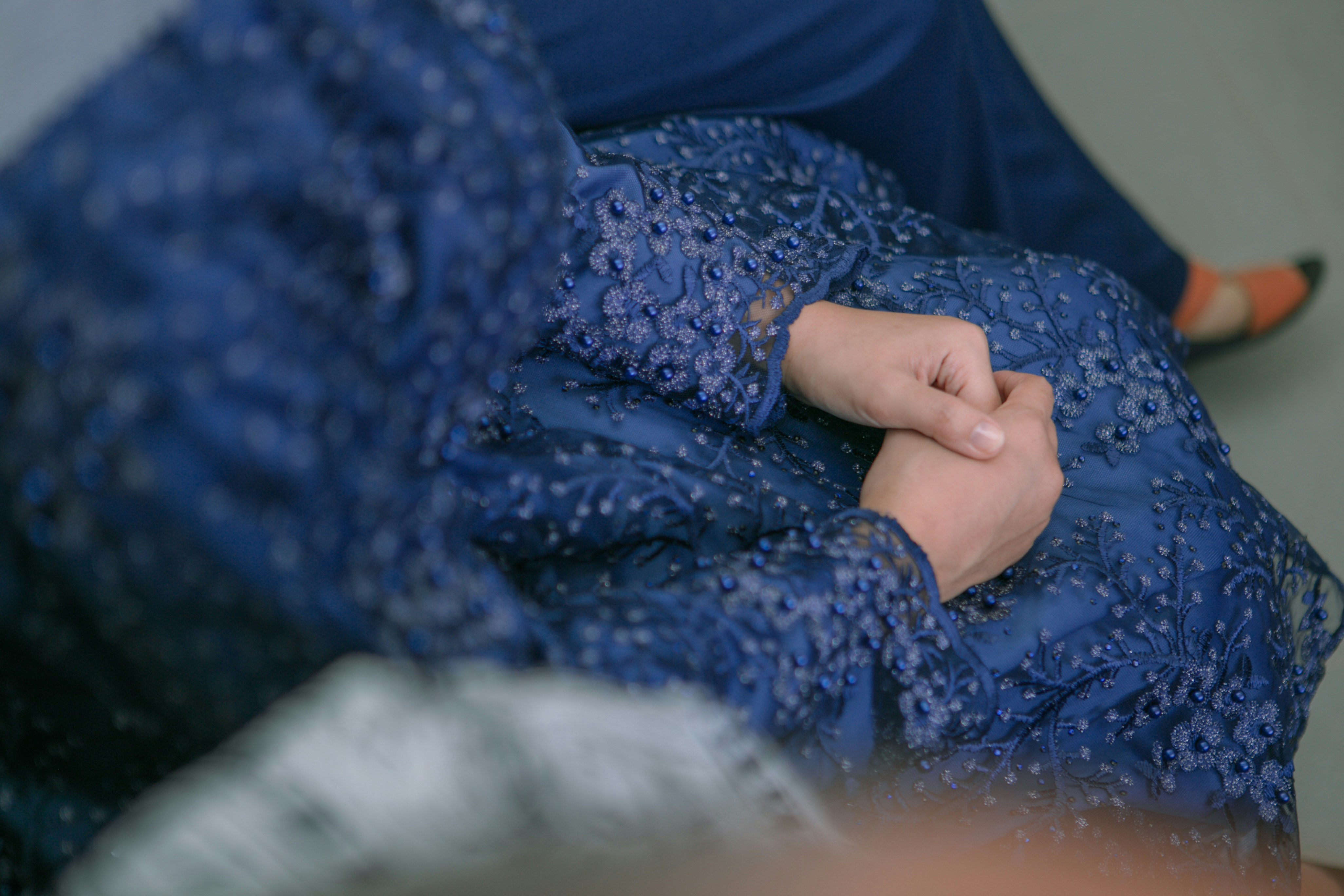 Intricately embroidered blue fabric drapes over clasped hands, showcasing a moment of contemplation. The soft focus highlights the texture and detail of the attire.