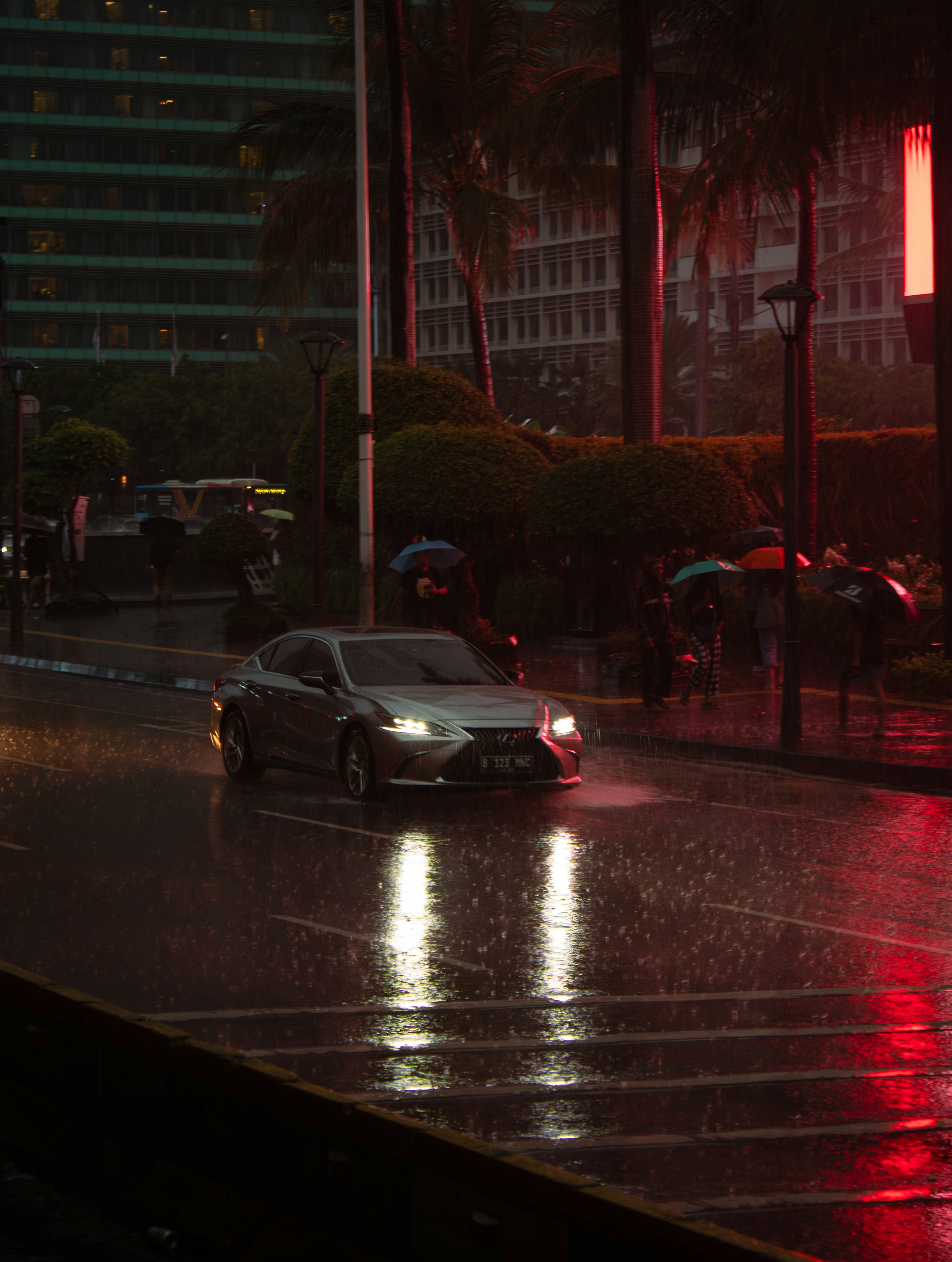 A sleek car navigates a rain-soaked street, reflecting vibrant city lights amidst a backdrop of umbrellas and palm trees.