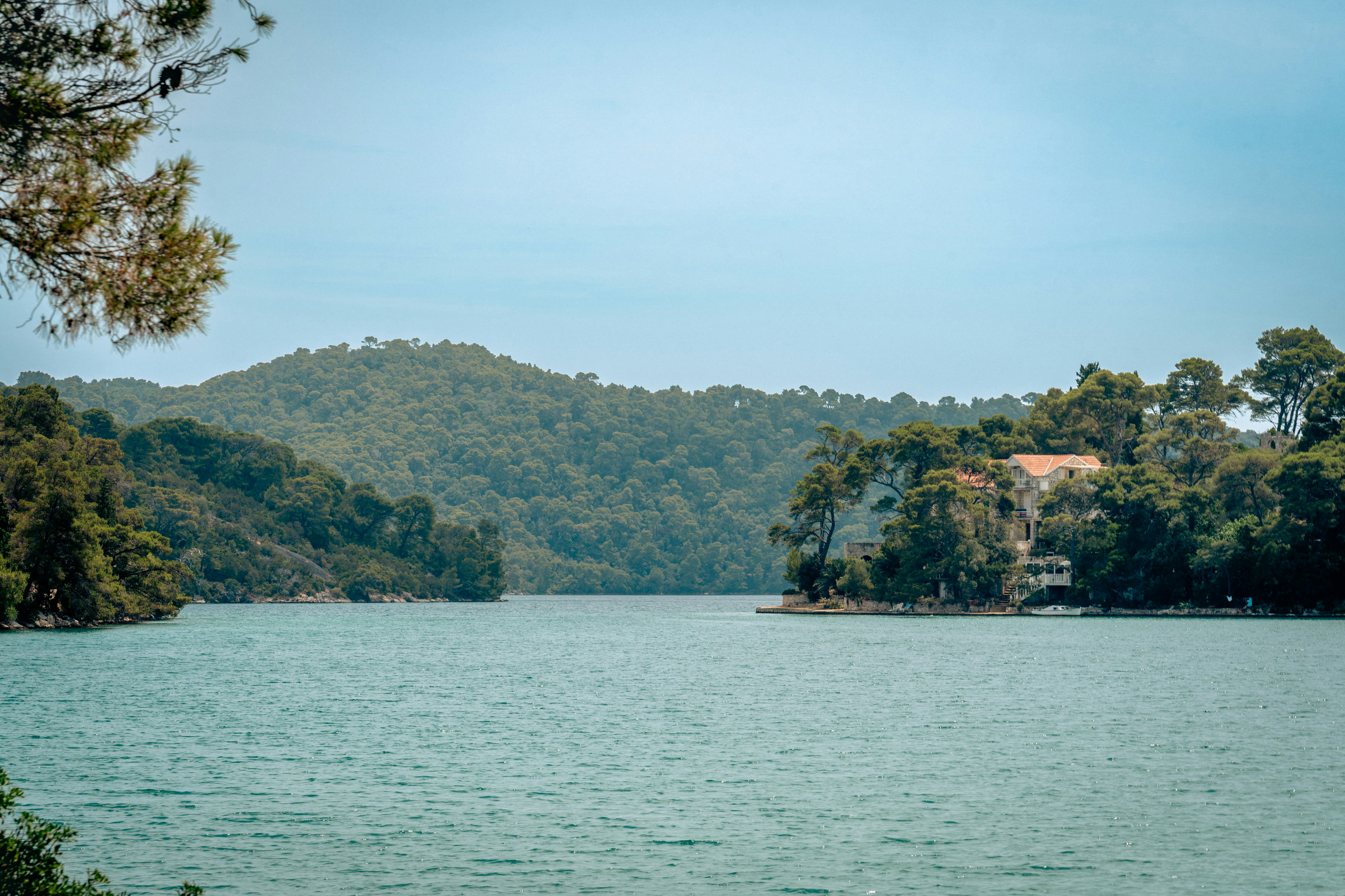 Tranquil lakeside view featuring lush greenery and a distant hillside with a charming residence. The calm water reflects the surrounding landscape.