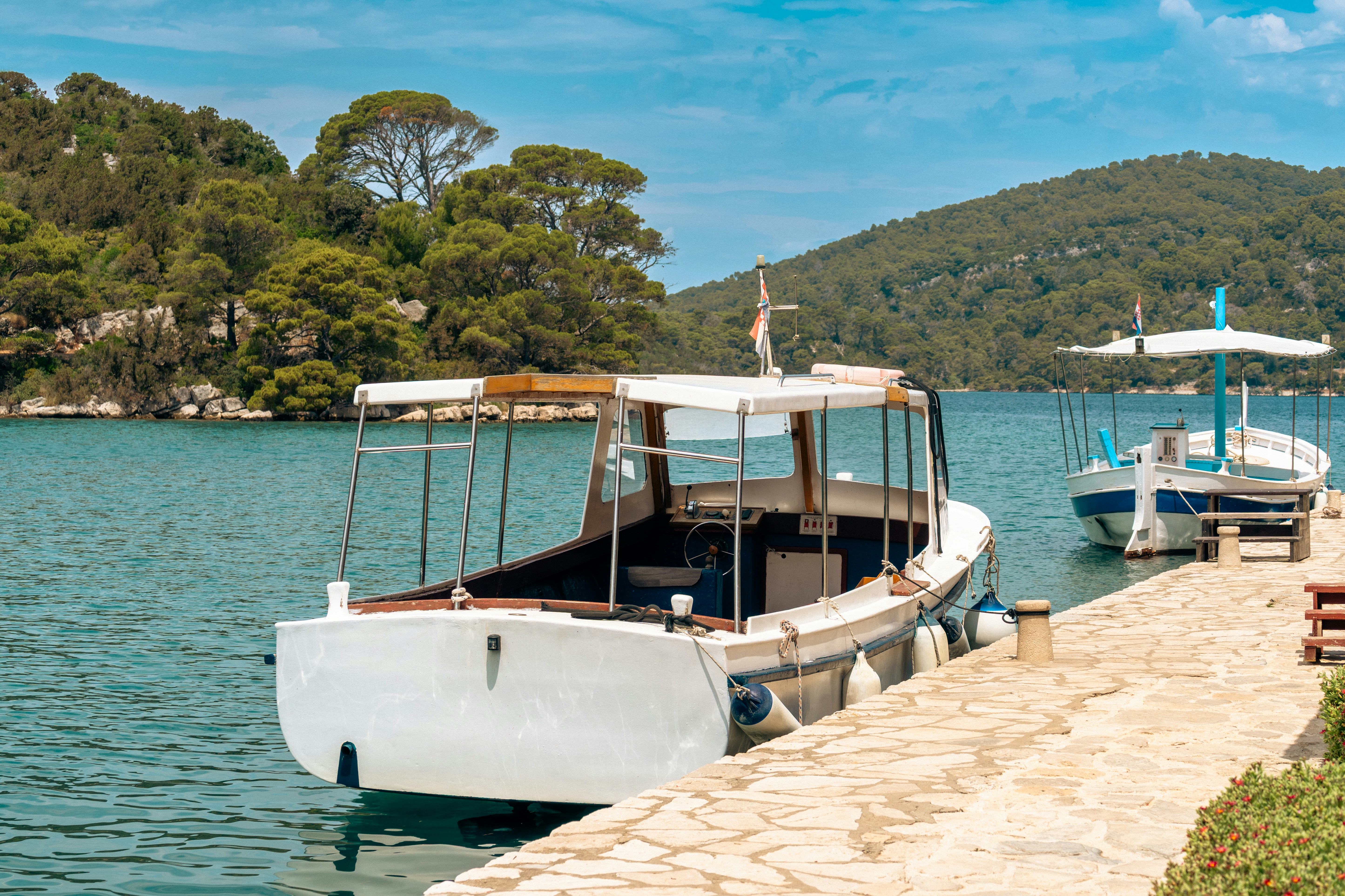 A classic boat moored at a stone dock, surrounded by lush greenery and calm waters under a clear blue sky.