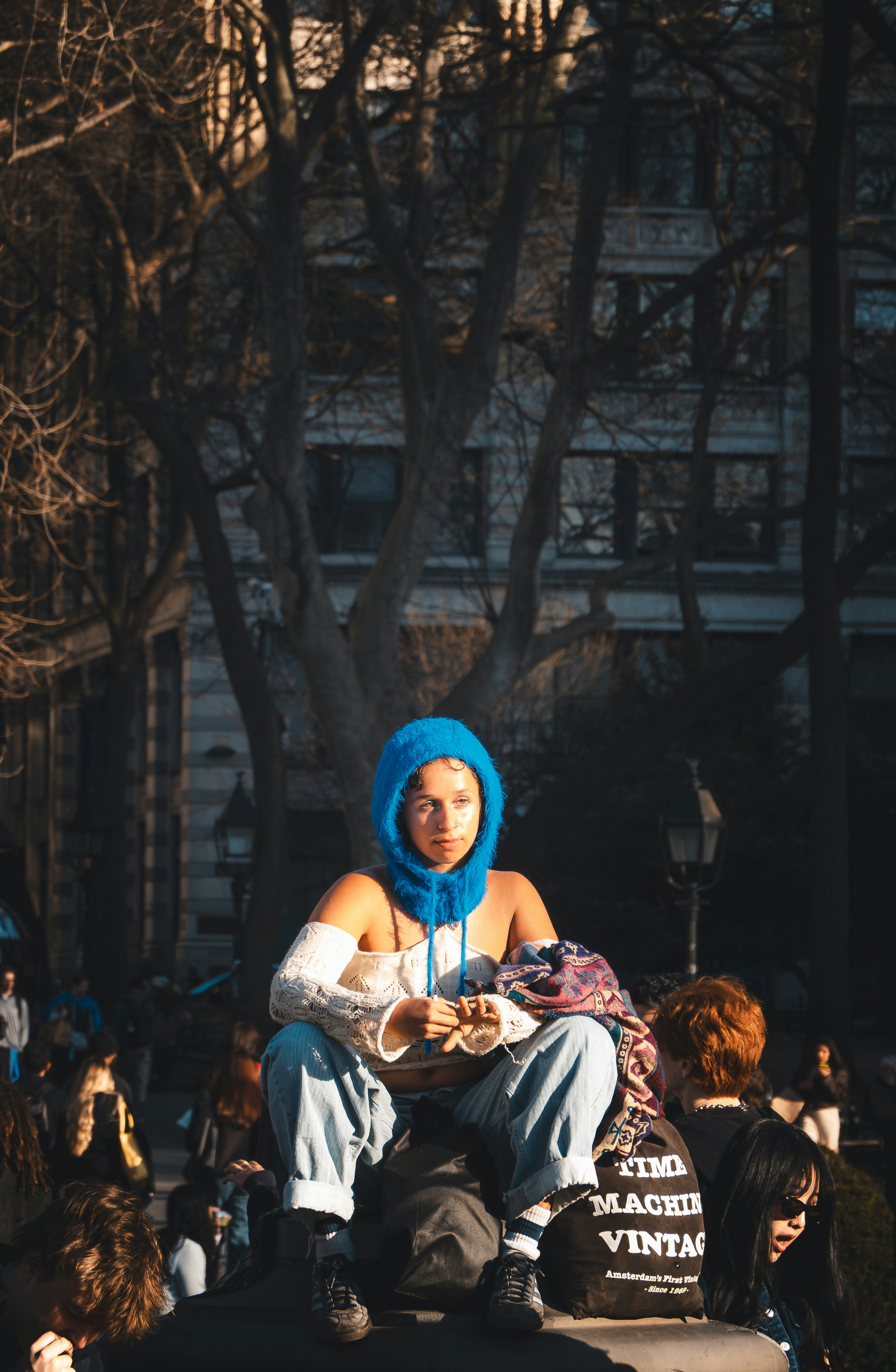 Young woman in a blue hood sitting on a statue, surrounded by a lively crowd in an urban park. Sunlight casts a warm glow on the scene.