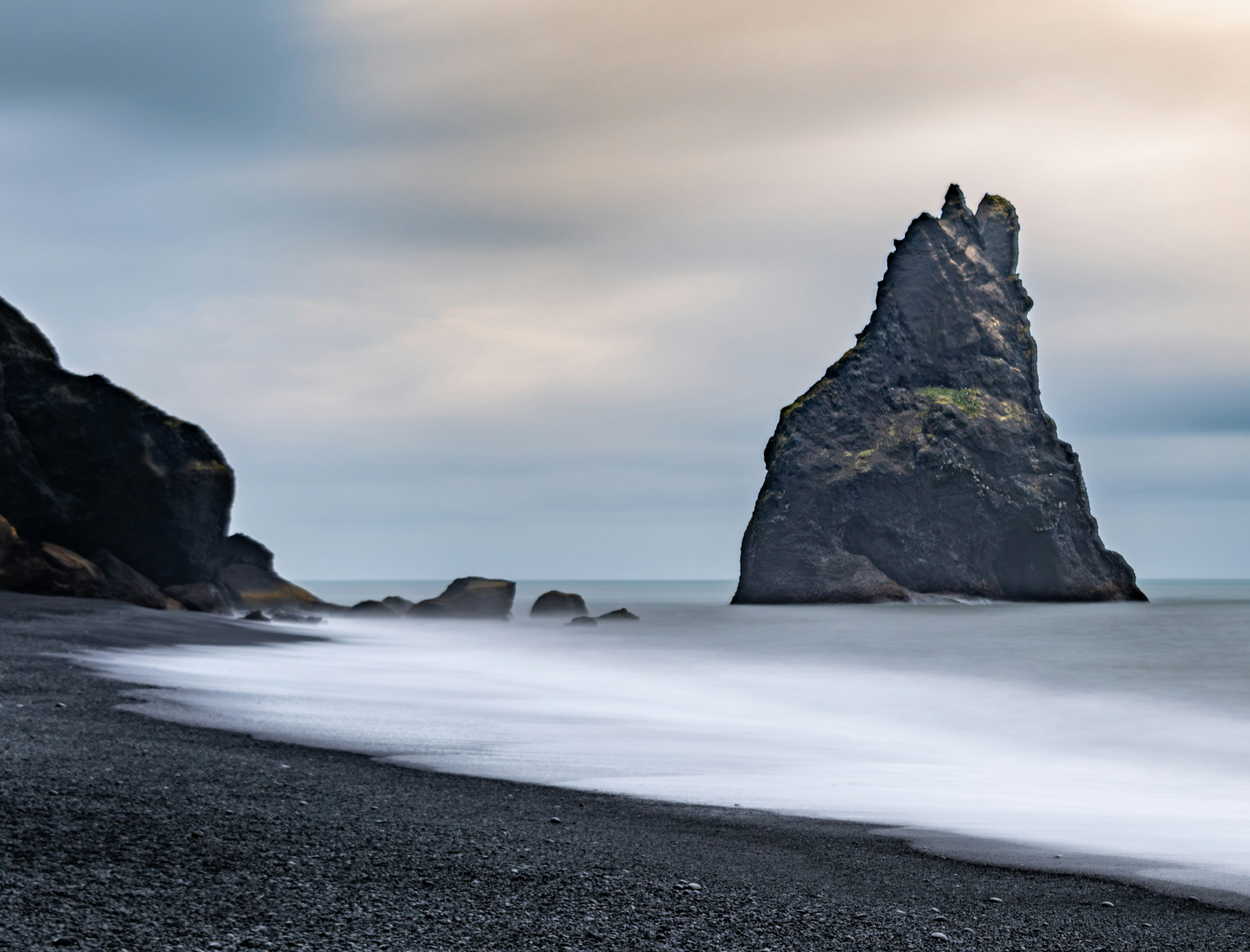 A dramatic sea stack rises from the calm waters, surrounded by a smooth black sand beach under a moody sky.