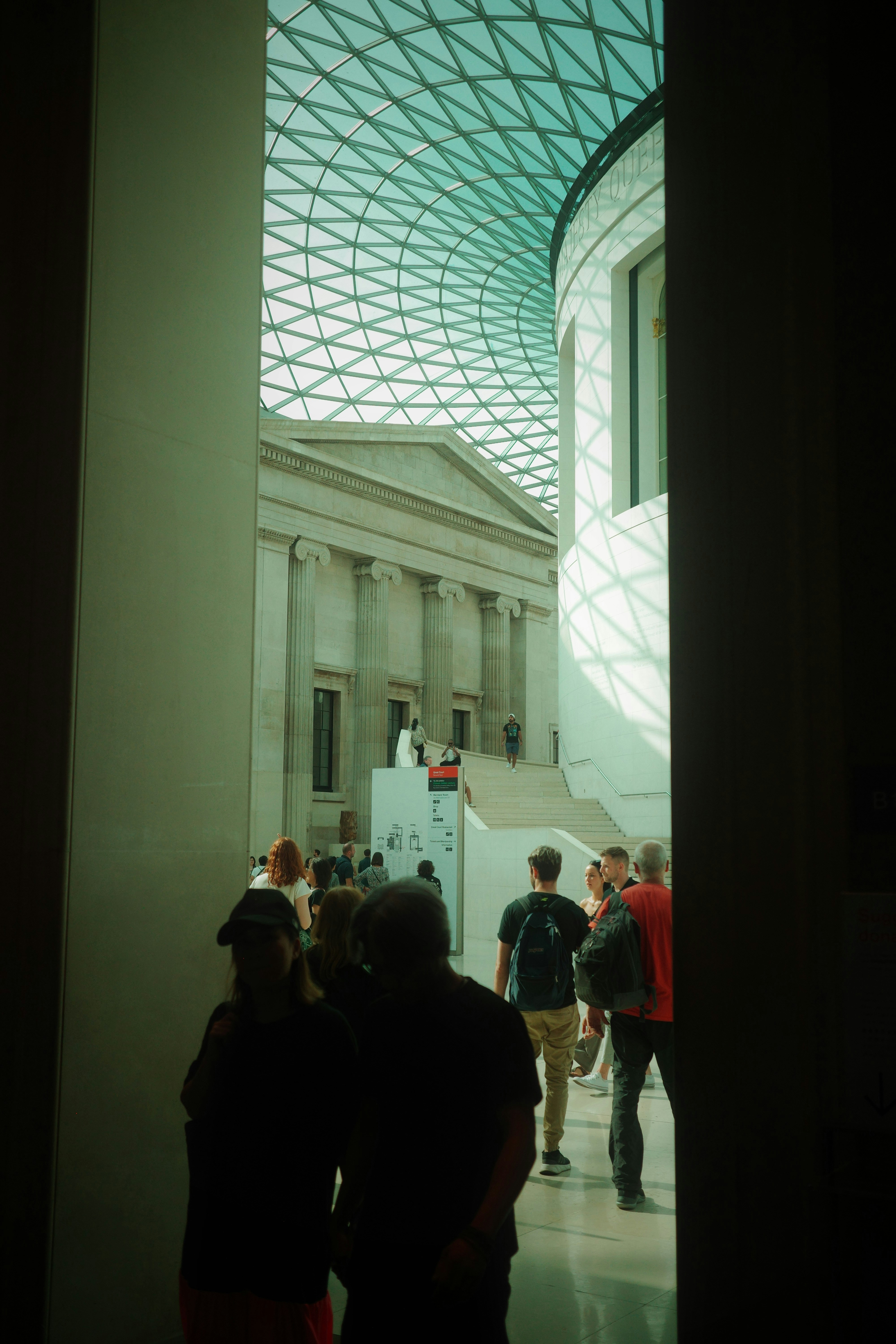 Visitors exploring the British Museum, framed by the modern glass ceiling and classical architecture. The interplay of light and shadows enhances the viewing experience.