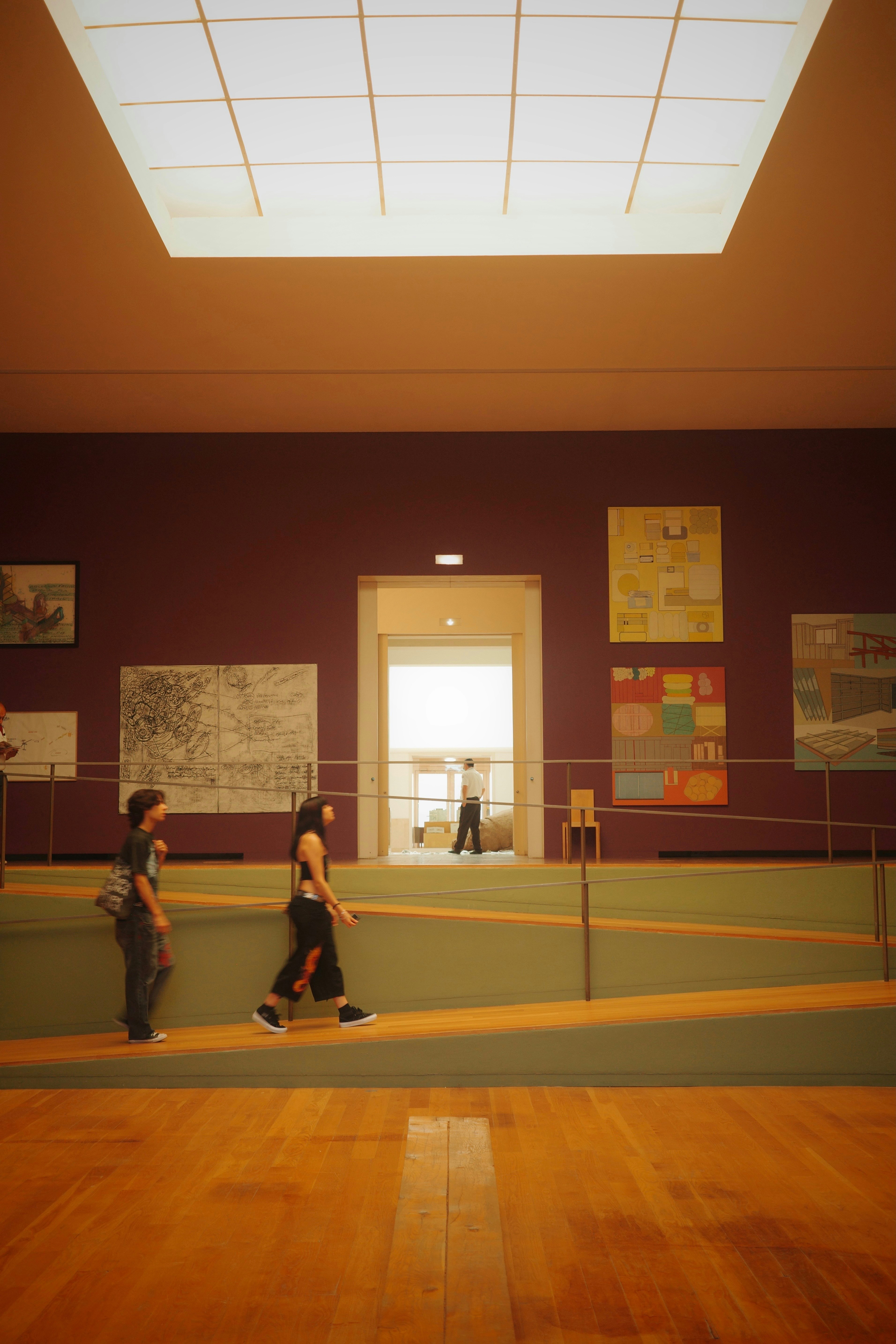 Visitors stroll through a modern art gallery, framed by vibrant wall art and a luminous ceiling. The scene captures the blend of human interaction and artistic expression.