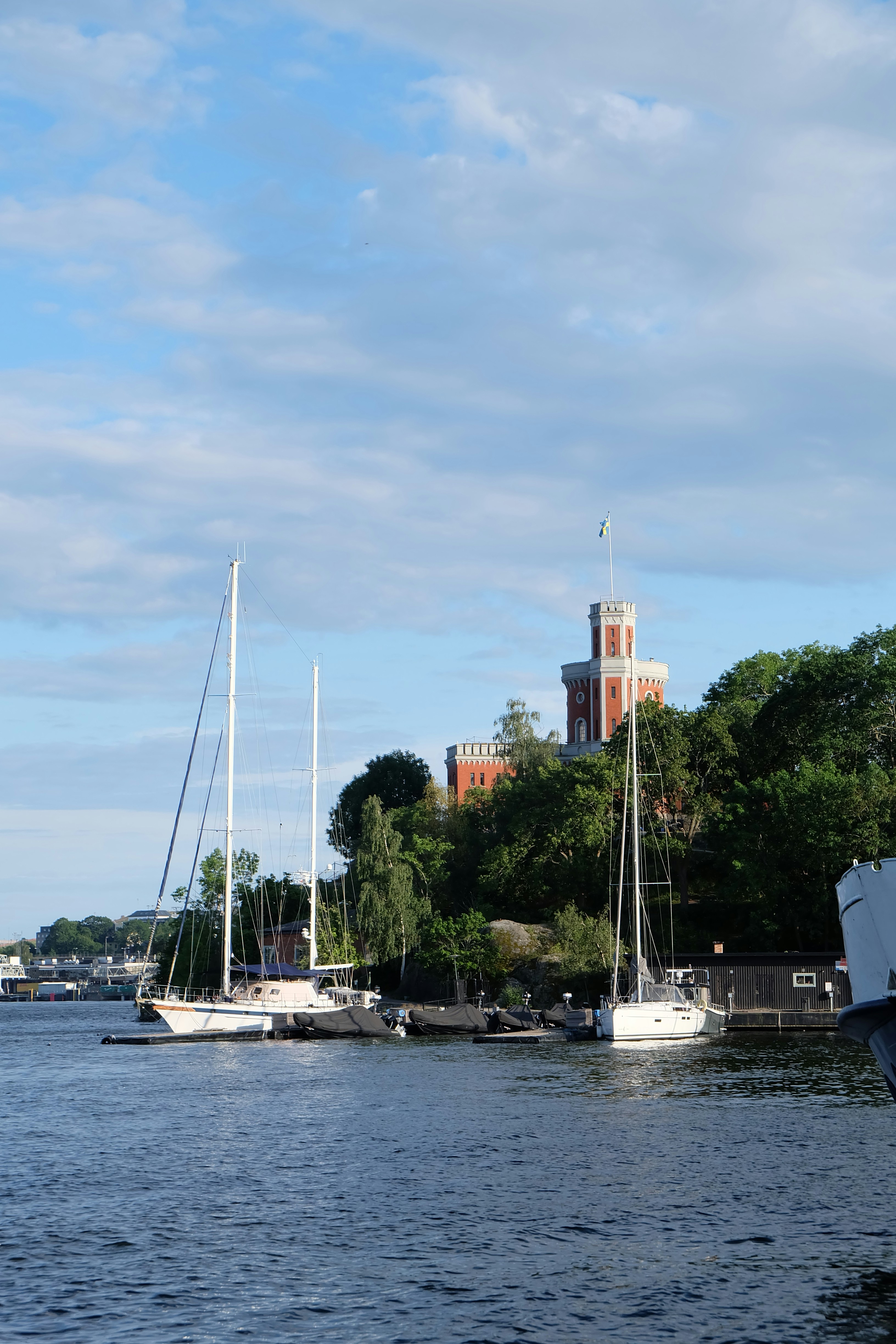 Sailboats anchored in a tranquil harbor, with a historic tower peeking through lush greenery under a partly cloudy sky.