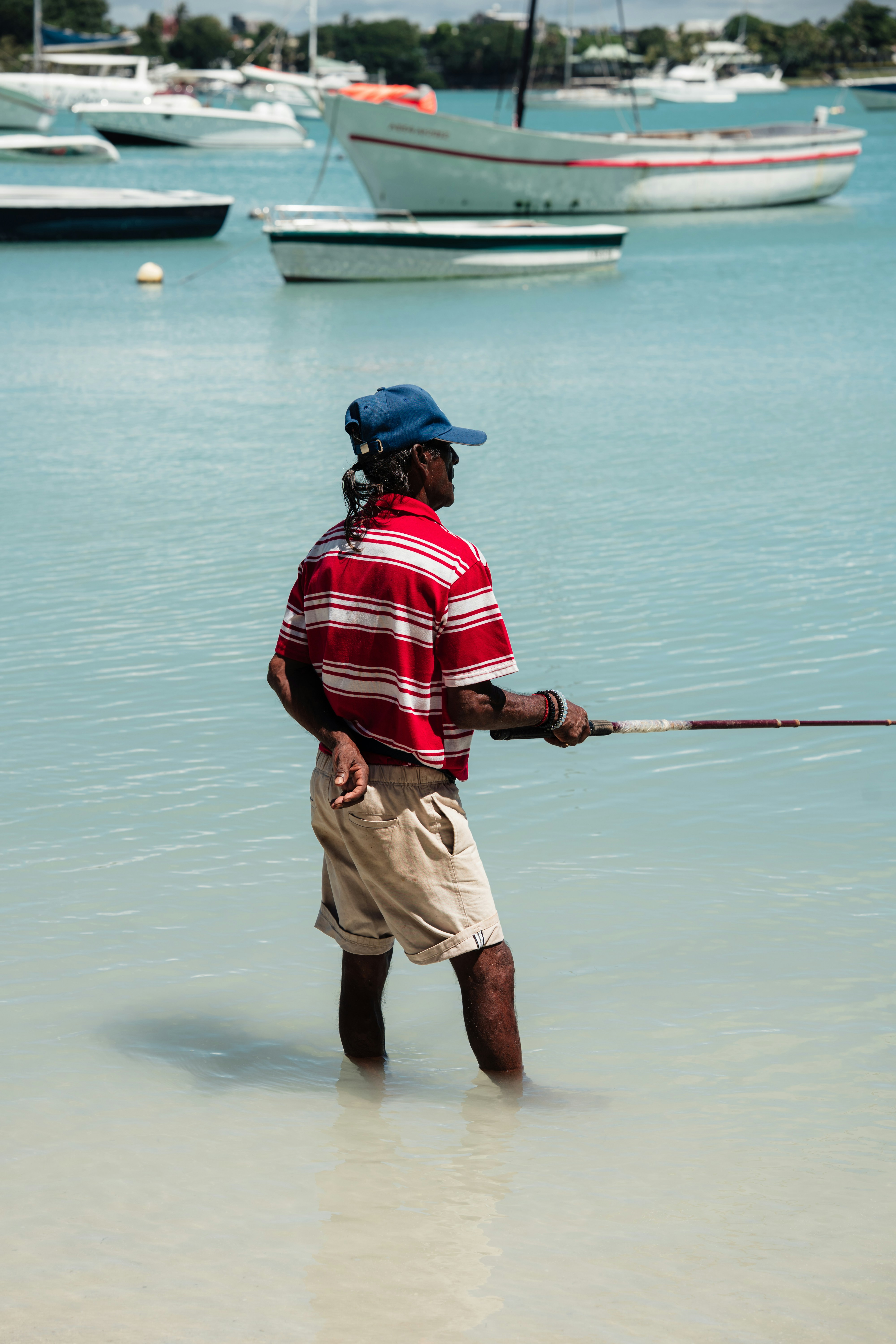 A man fishes in shallow water near boats.