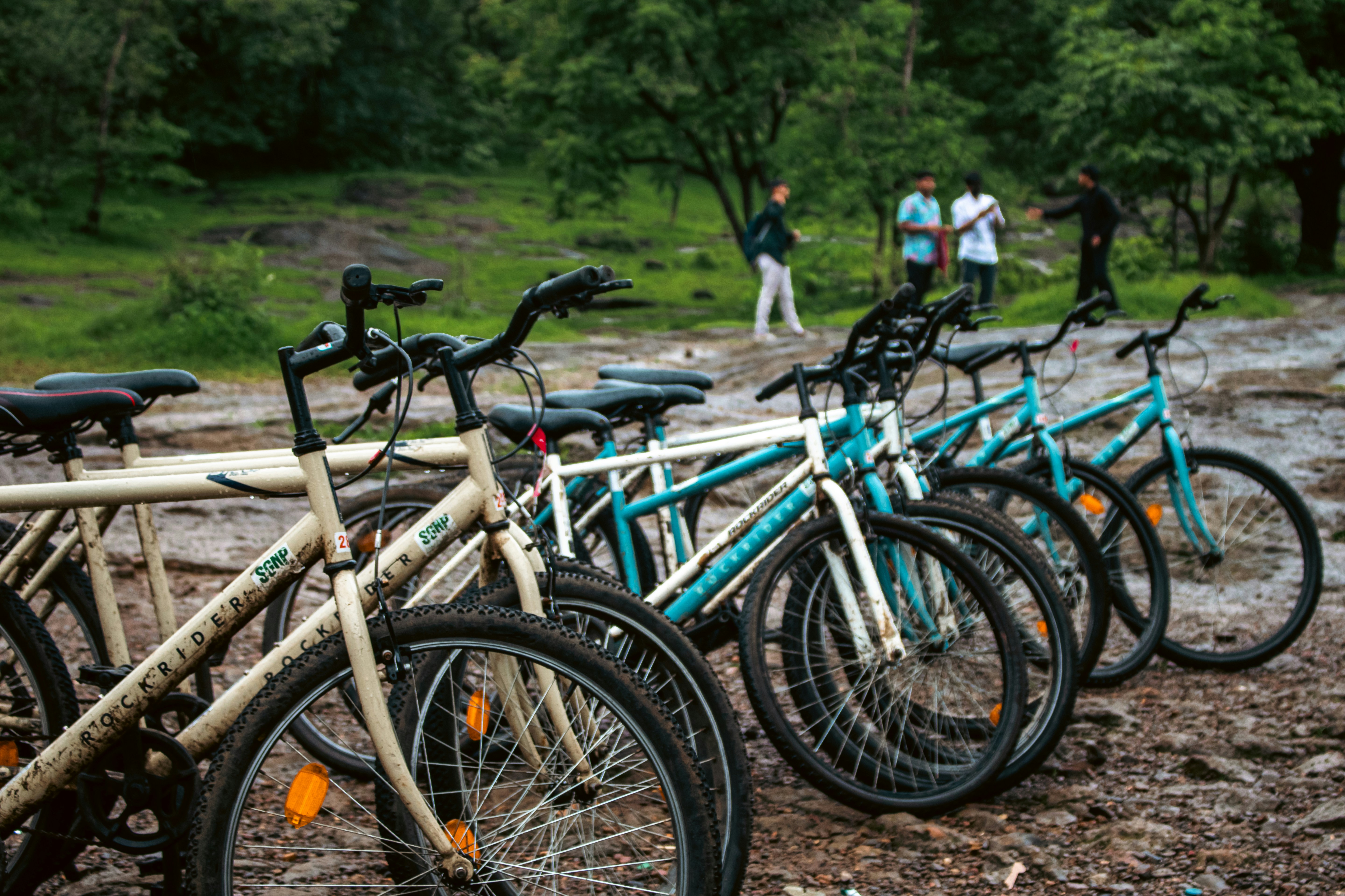 A group of trekking bicycles with rugged terrain tires are parked on a muddy patch of ground, after an adventurous ride through off-road trails or forest paths. | Bicycles lined up with people in the background.