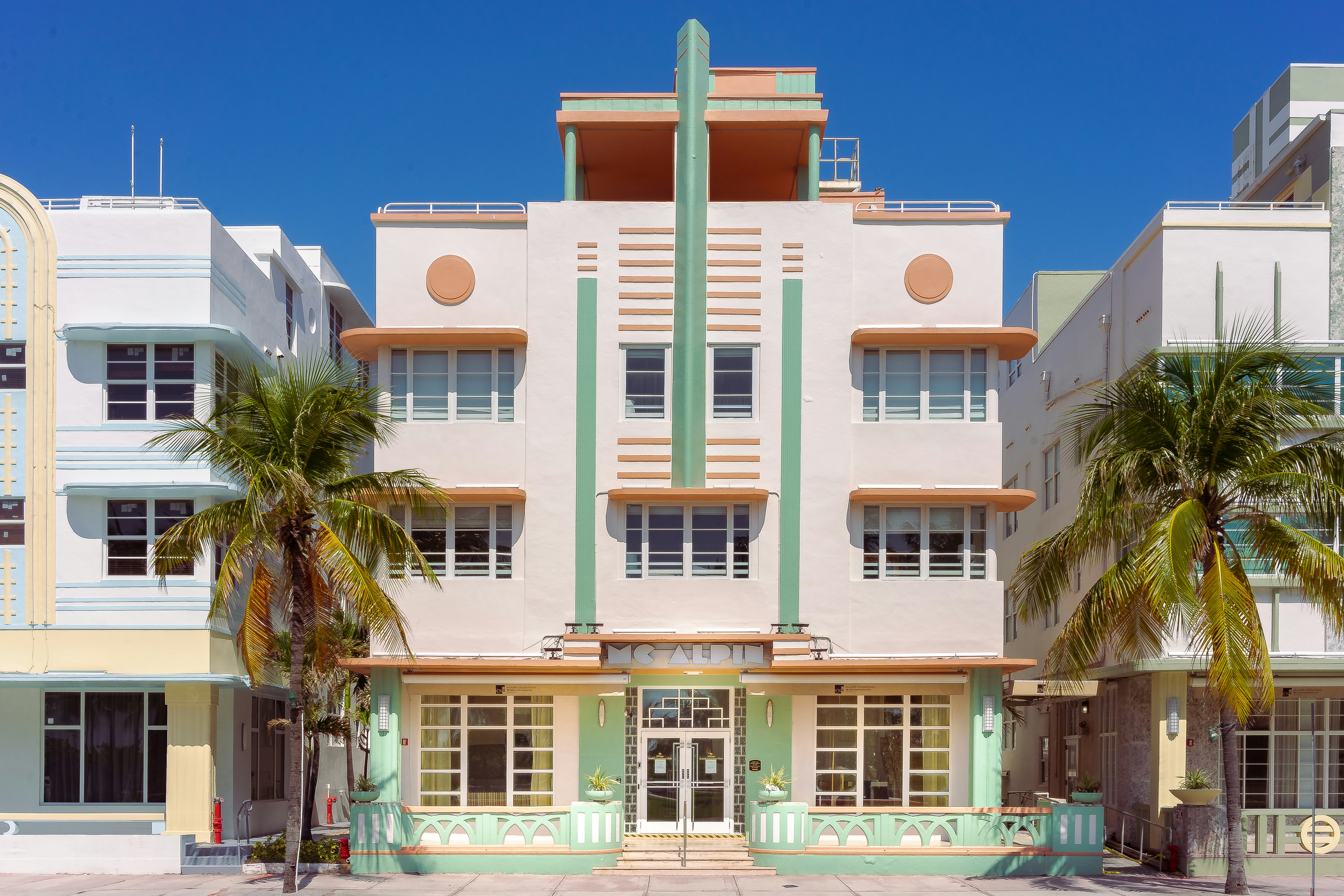 A pastel-hued Art Deco building in Miami’s South Beach stands tall under a vibrant blue sky, flanked by palm trees. Its symmetrical façade and retro colors evoke the charm of vintage Florida architecture. | Art deco architecture in miami beach.