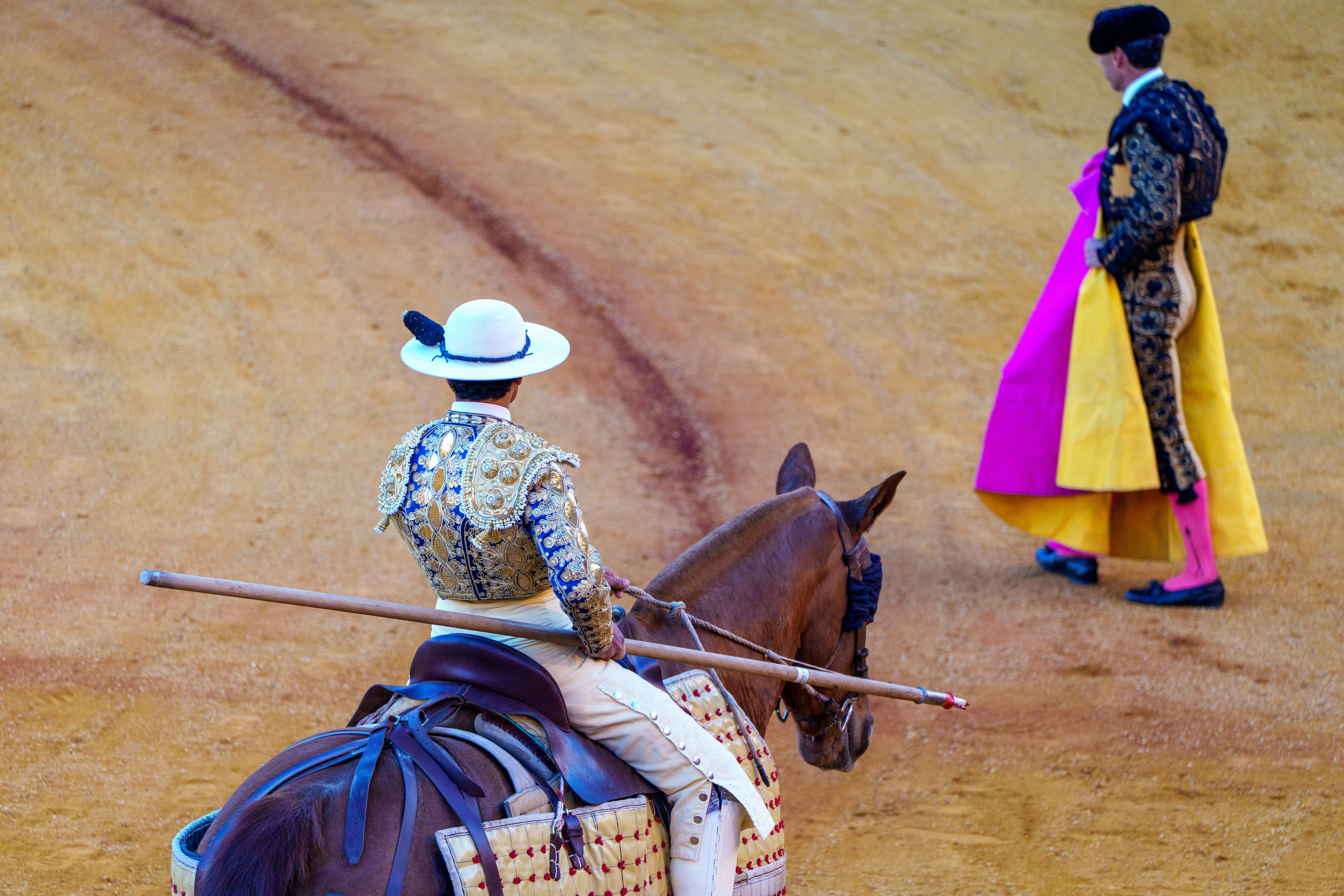A matador on horseback prepares for the duel, showcasing intricate attire, while another matador stands poised in vibrant colors, set against the sandy arena backdrop.